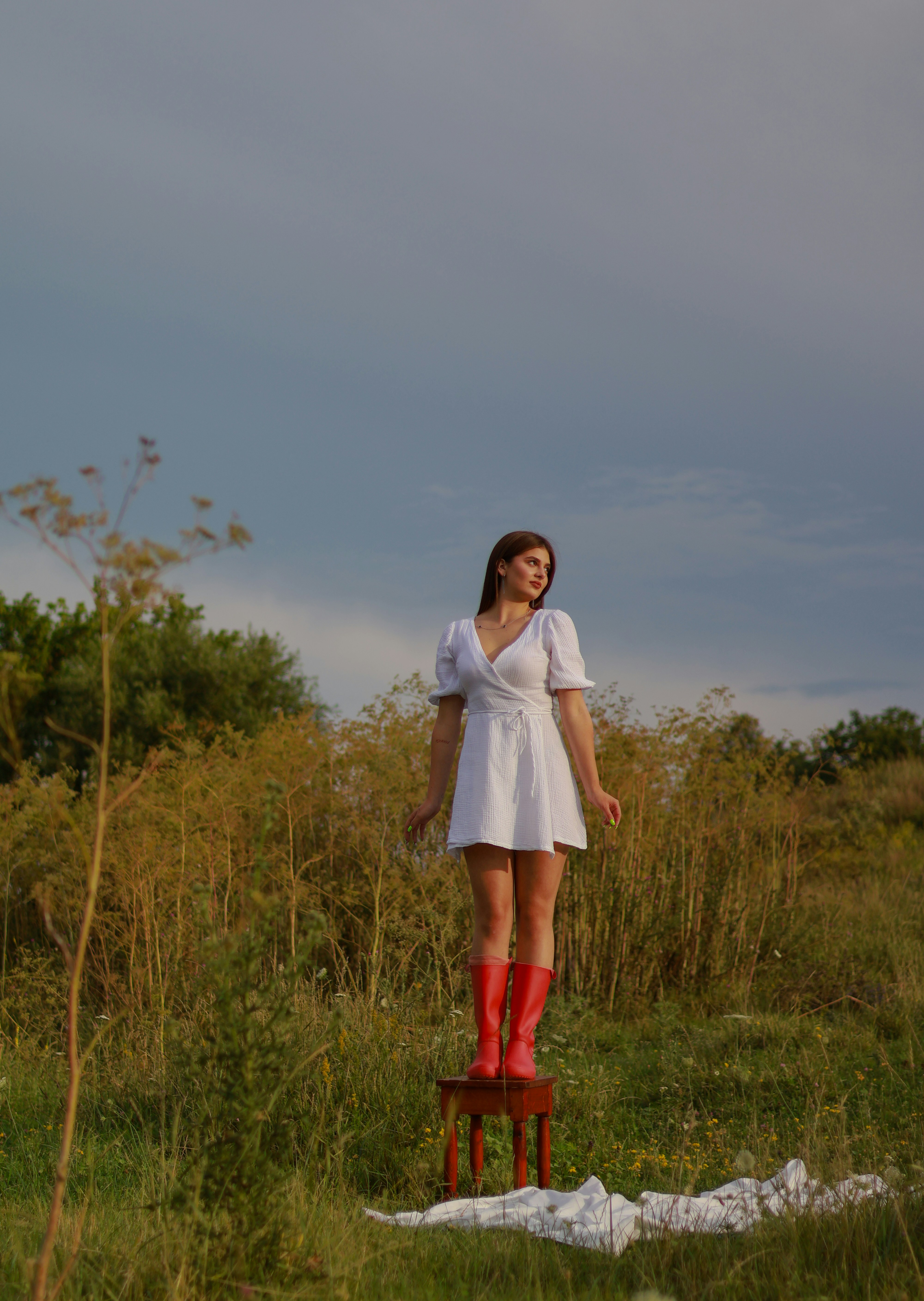 Photograph of a woman in a white dress standing on a small wooden stool in a sunlit meadow, wearing knee-high red boots. A white fabric lies on the ground nearby, with an open blue sky backdrop.
