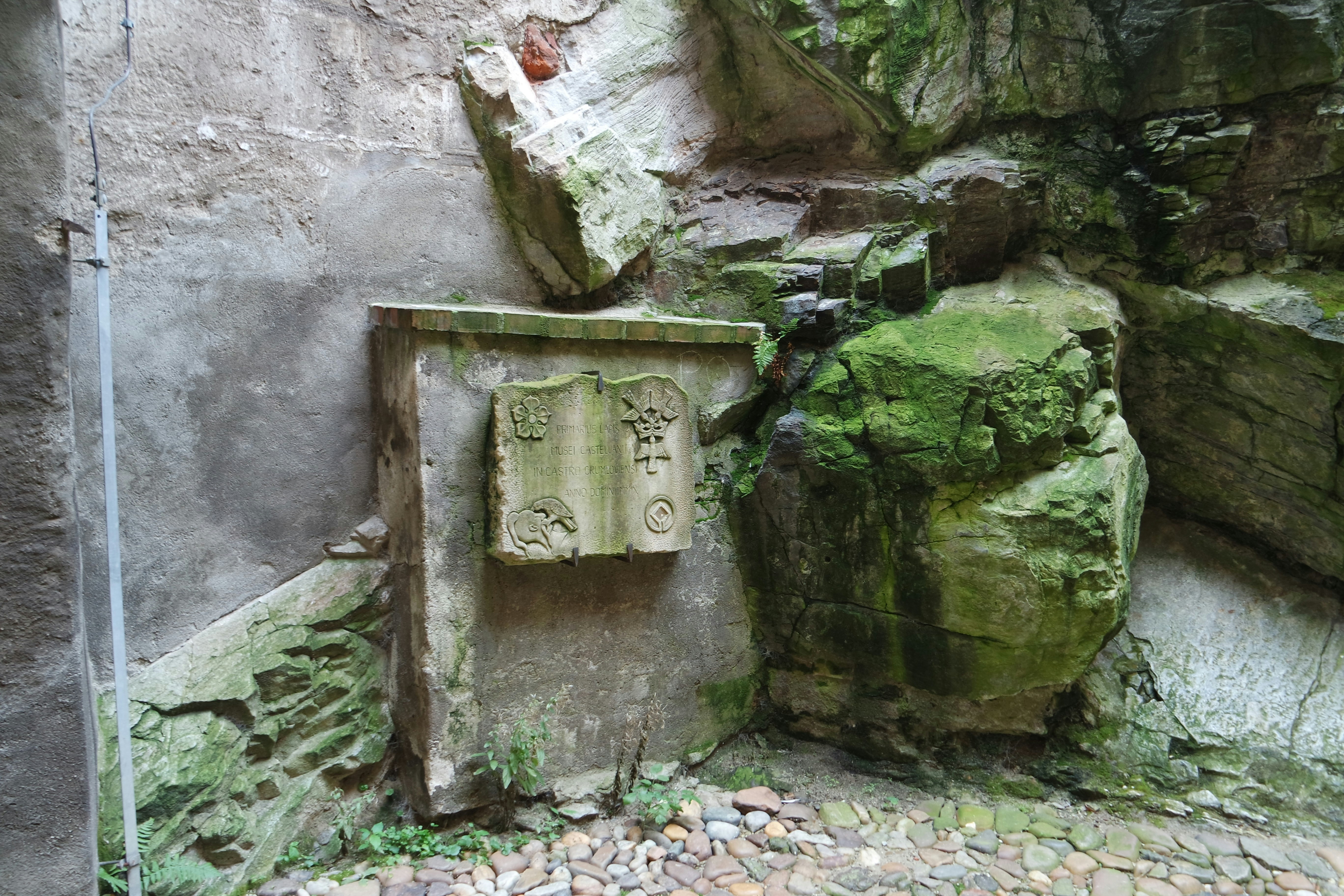 Weathered stone reliefs adorned with symbols, nestled against a moss-covered rock wall in a secluded alley.