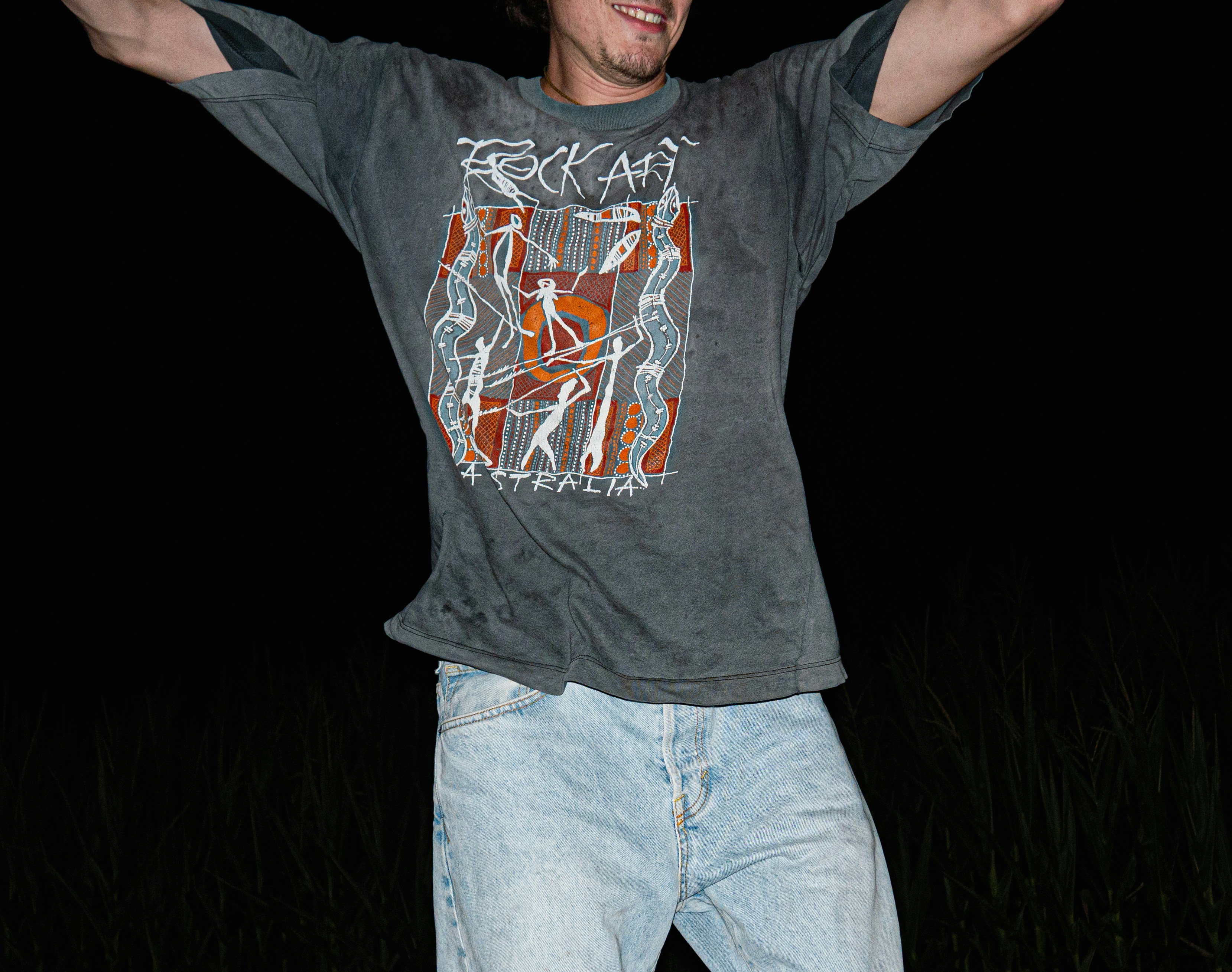 Man wearing a dark T-shirt with a large graphic print raises his arms against a black night backdrop.