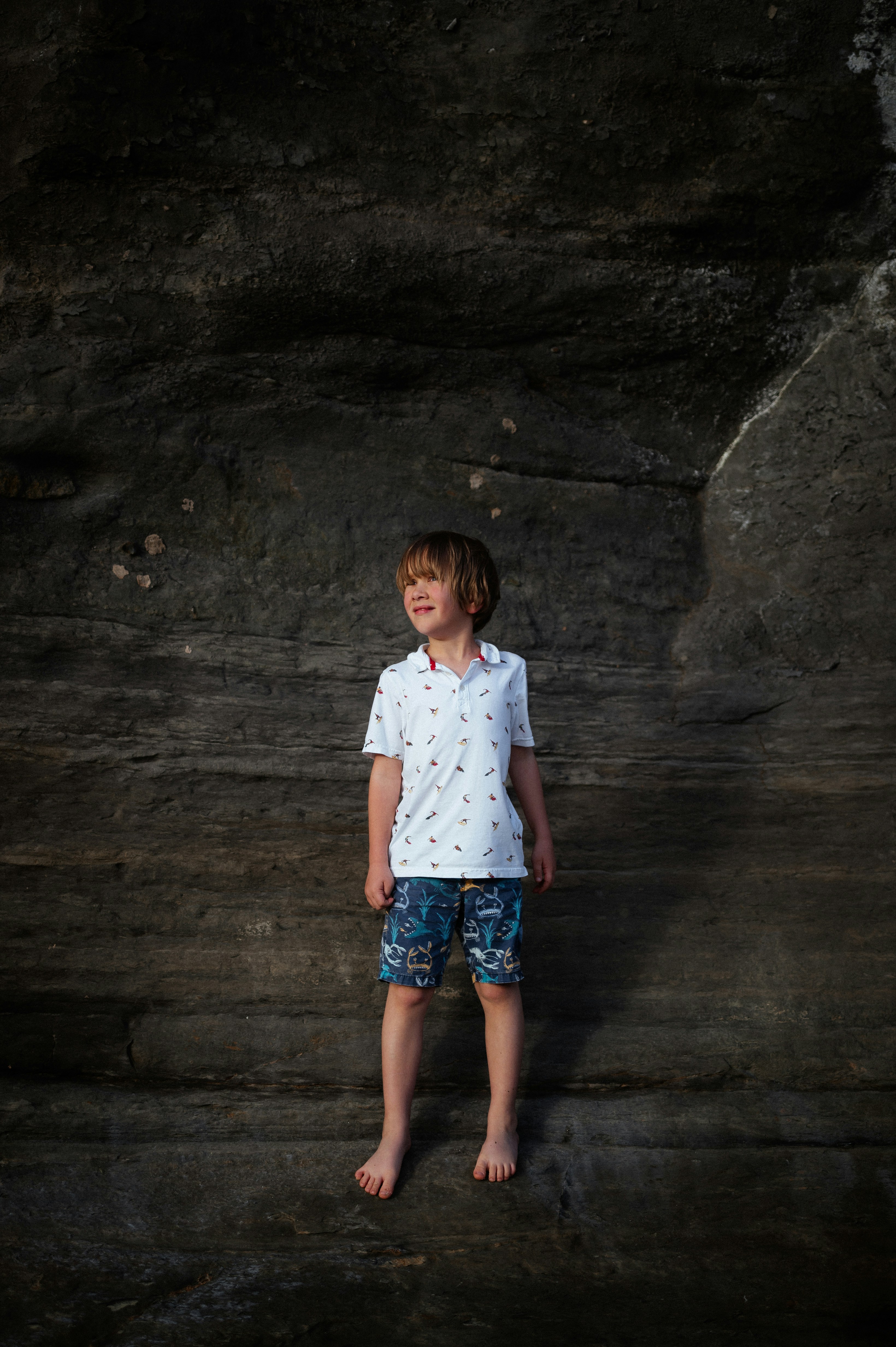 Barefoot boy in a white polo shirt stands against a layered rock face, bathed in soft side light.