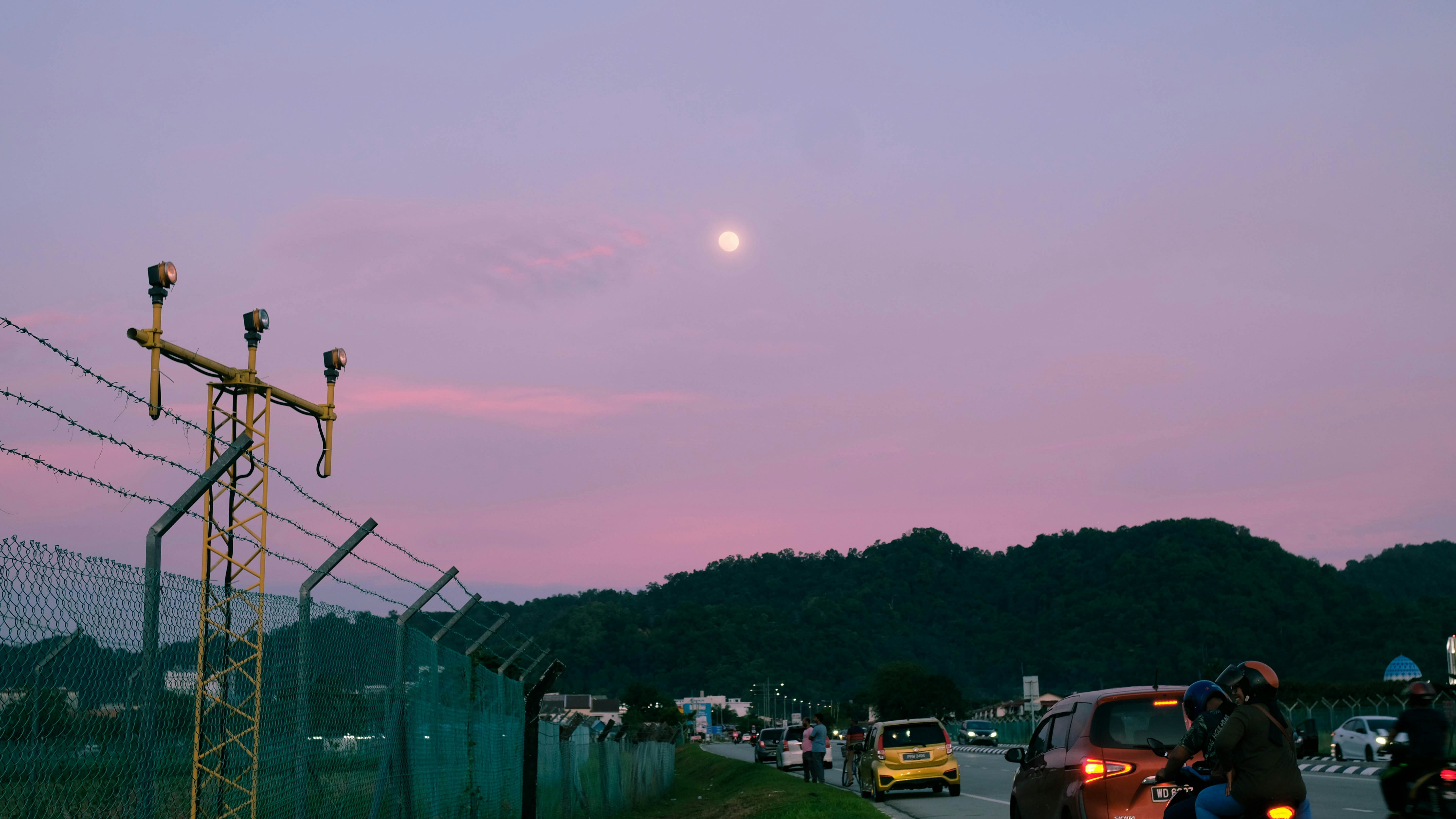 A serene twilight scene featuring a rising moon over a winding road, framed by a security fence and distant hills.