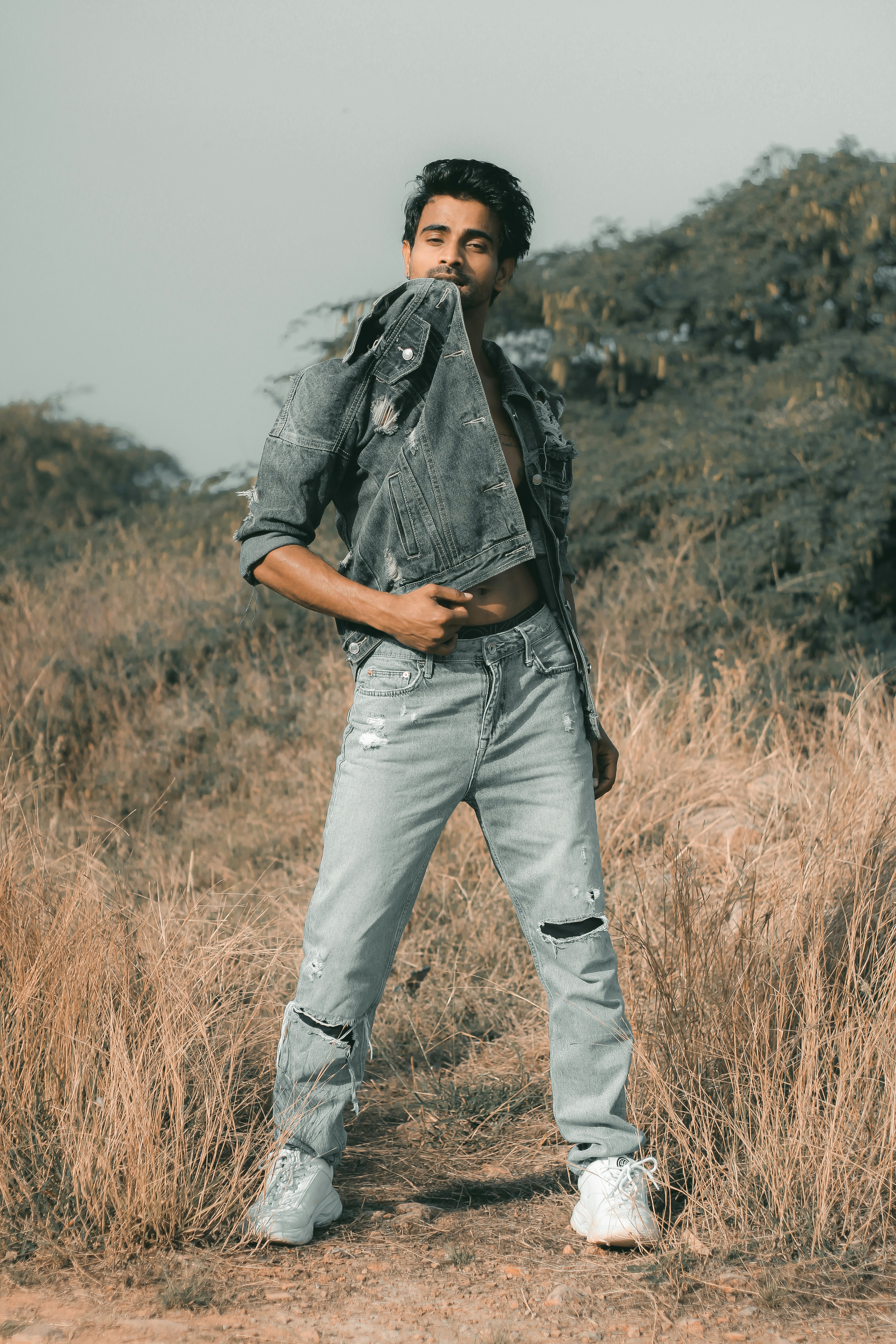 Male model stands in a sunlit, dry grass field, wearing a distressed denim jacket and jeans. He lifts the jacket to reveal his midriff in a bold, cinematic pose.