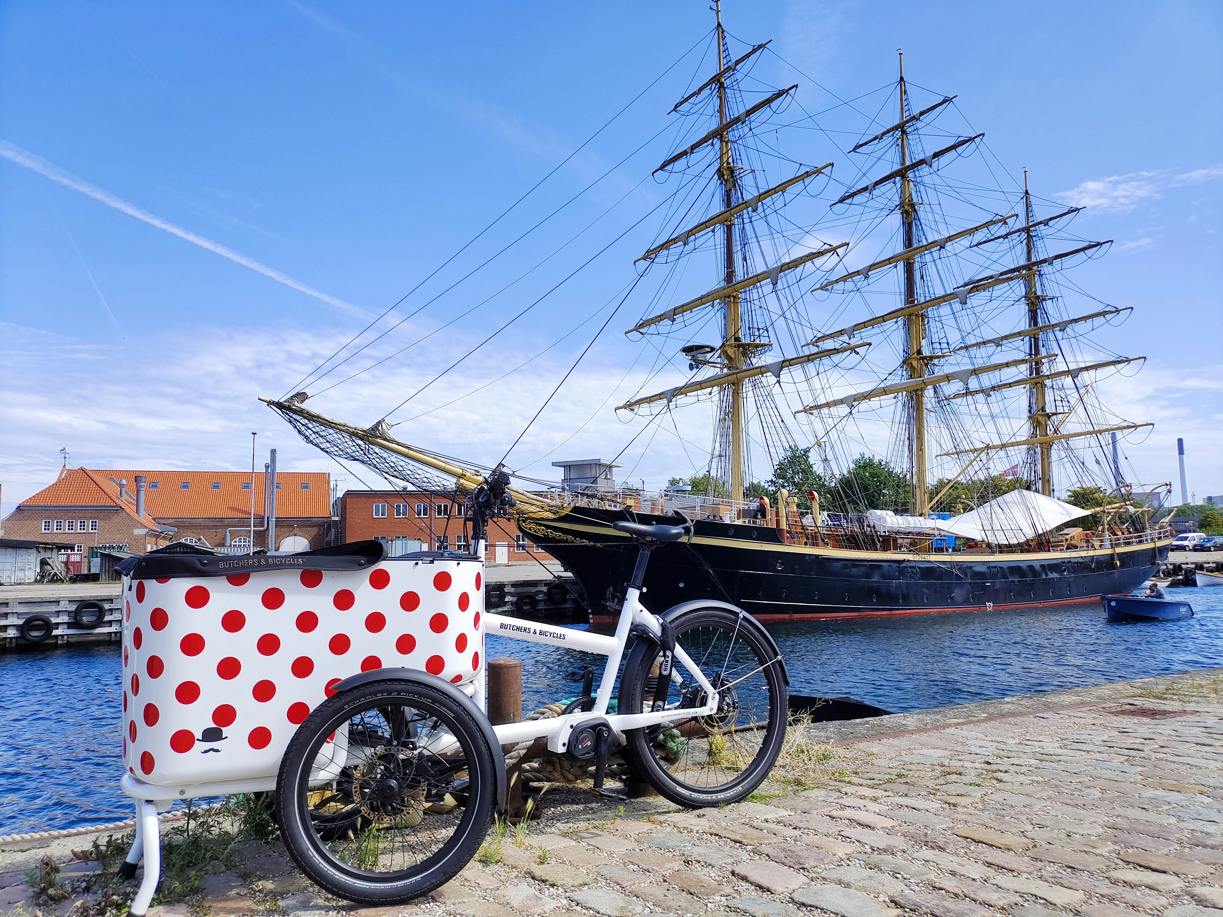 A white cargo bike with a red polka-dot box sits on a cobblestone quay, with a tall-masted ship moored in the blue harbor behind.
