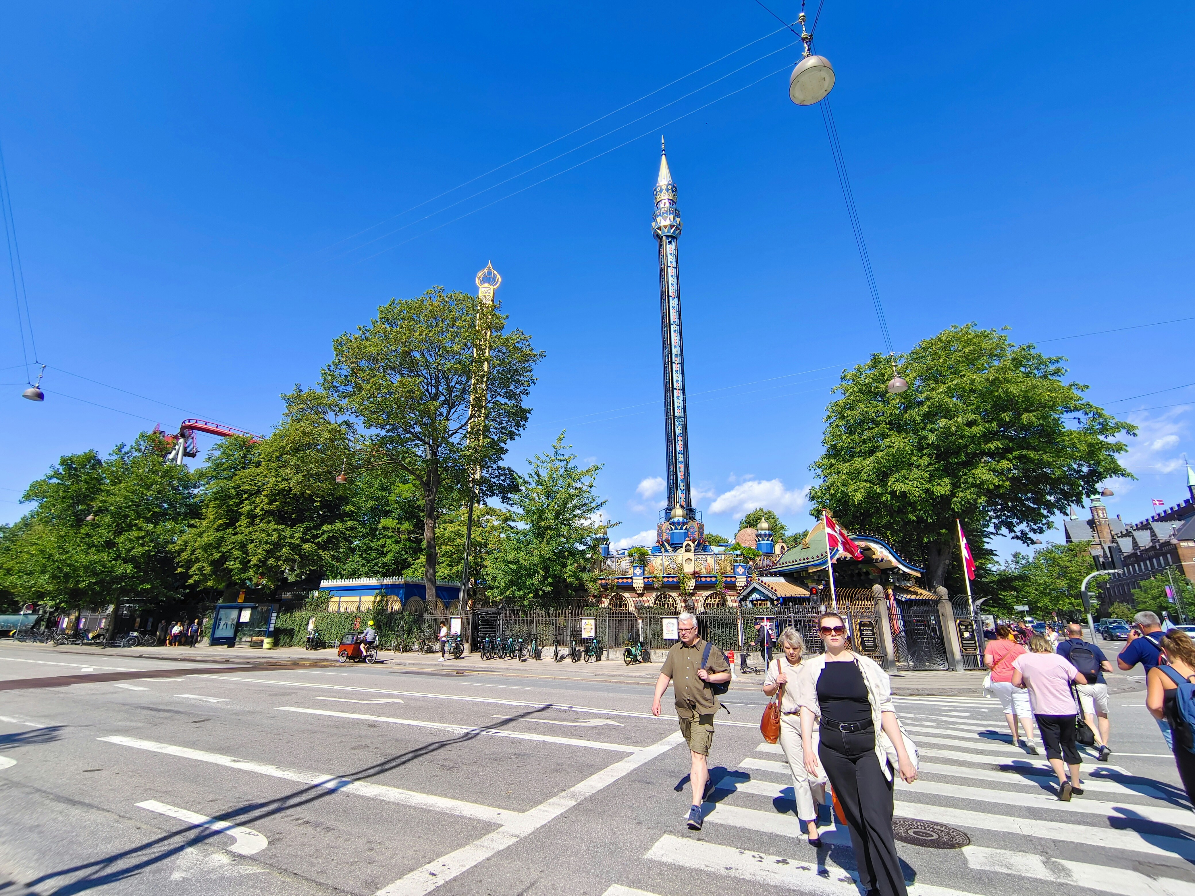 Sunlit city square featuring a tall clock-tower monument, surrounding trees, and pedestrians crossing a broad crosswalk.