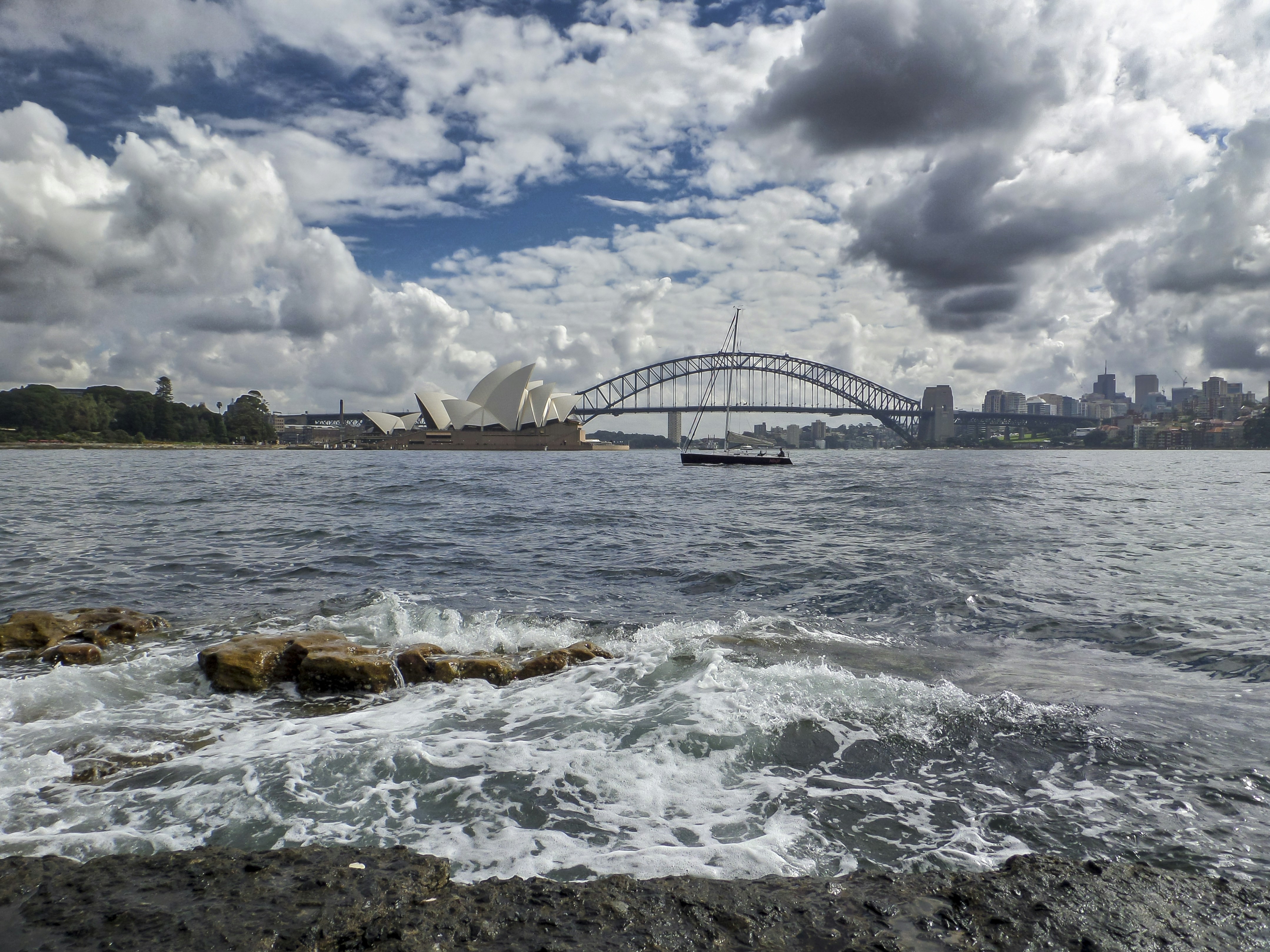 Harbor scene featuring the Sydney Opera House and Harbour Bridge across the water with rocks and waves in the foreground.