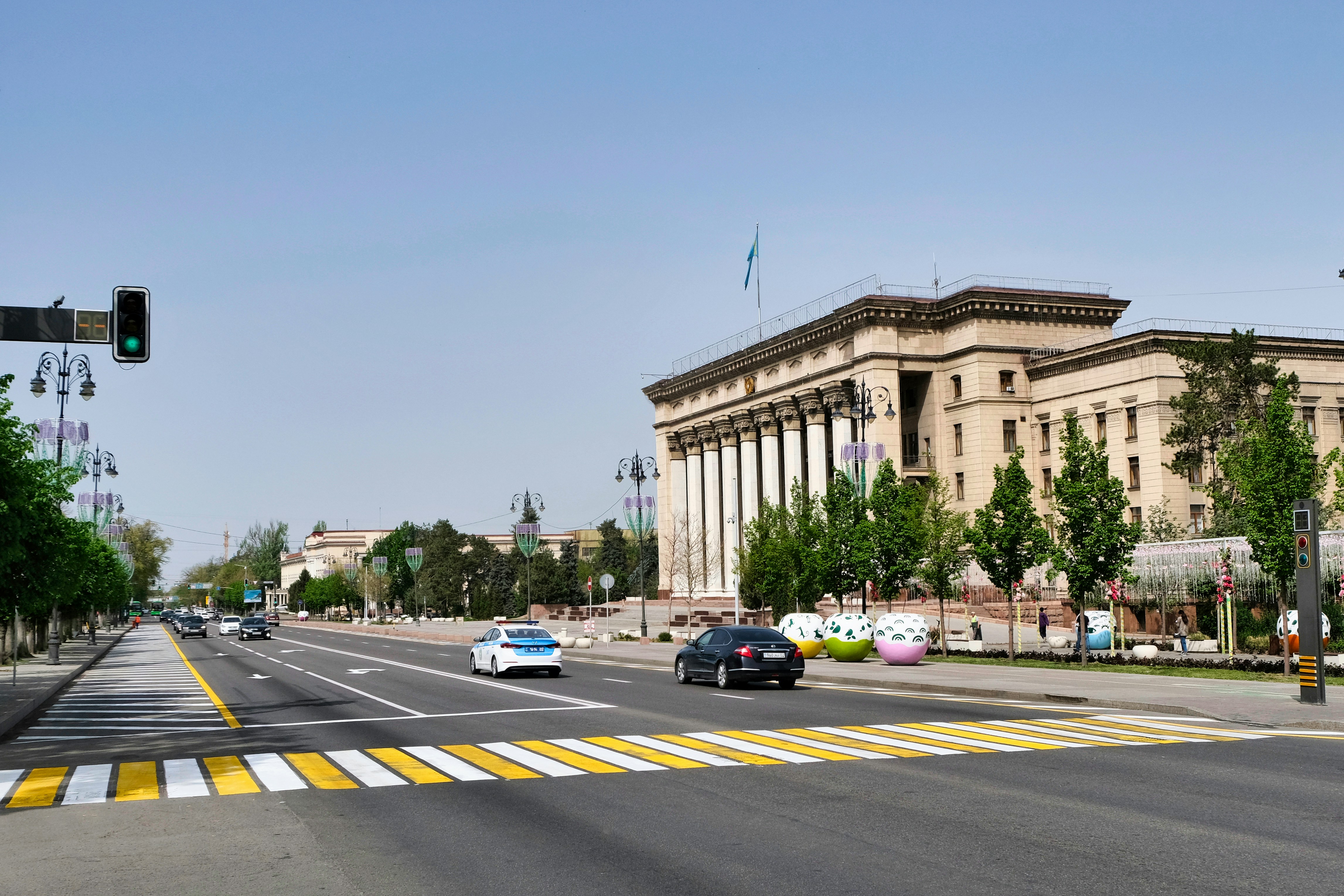 A bustling city street featuring a historic building with grand columns, flanked by trees and contemporary vehicles. The scene captures the blend of architectural styles in an urban environment.