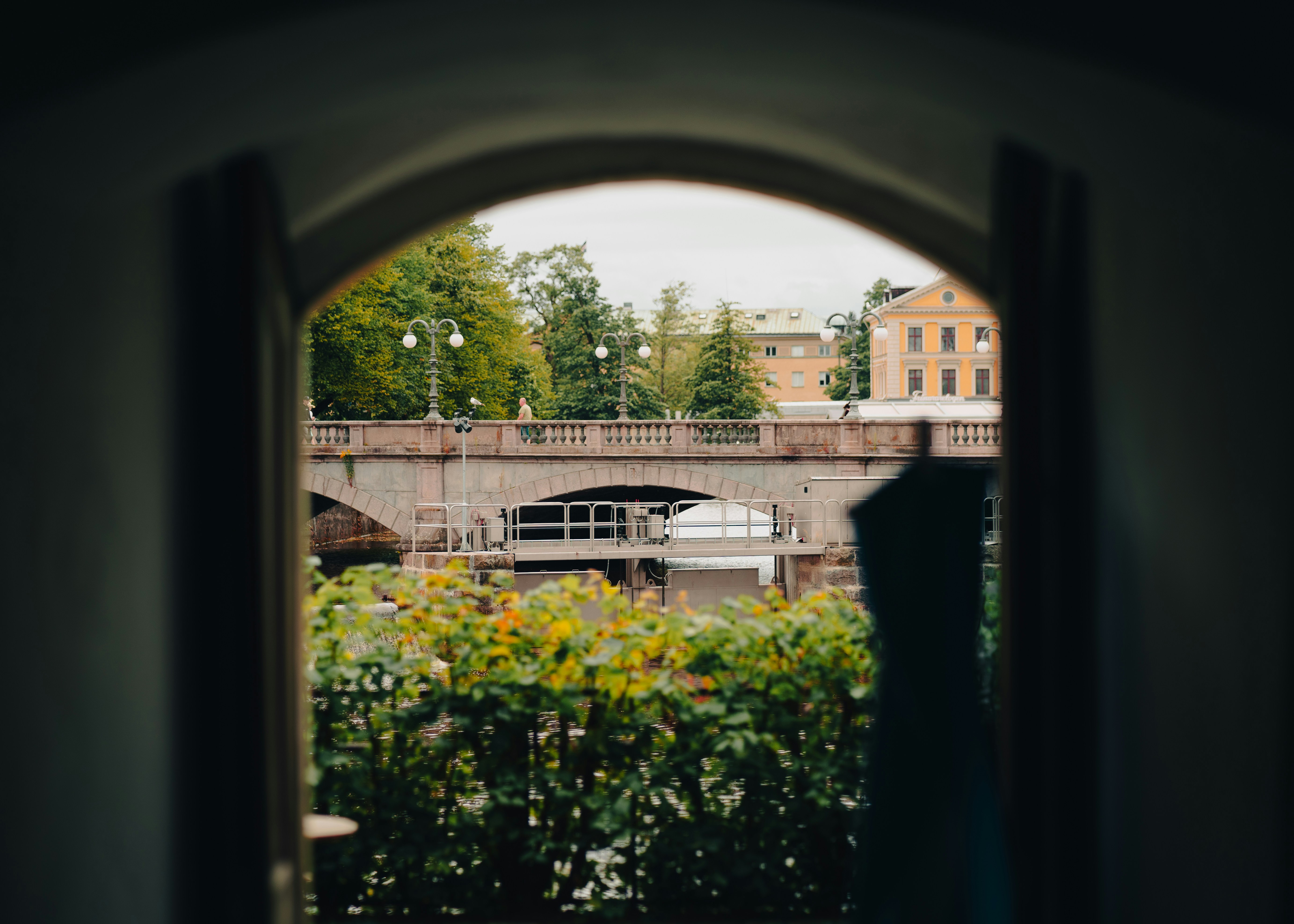 A view of a bridge framed by an archway, with lush greenery in the foreground and historical buildings in the background.