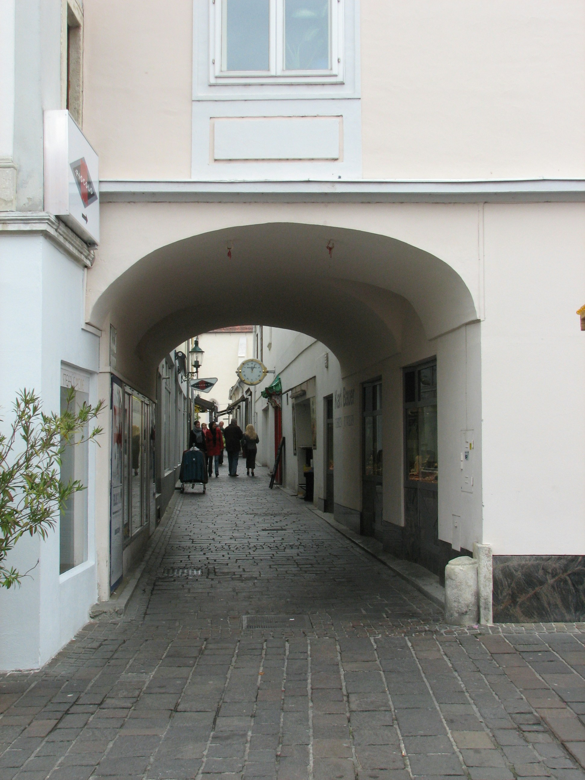 Photograph of a narrow cobblestone street seen through a white archway, with pedestrians walking in the distance.