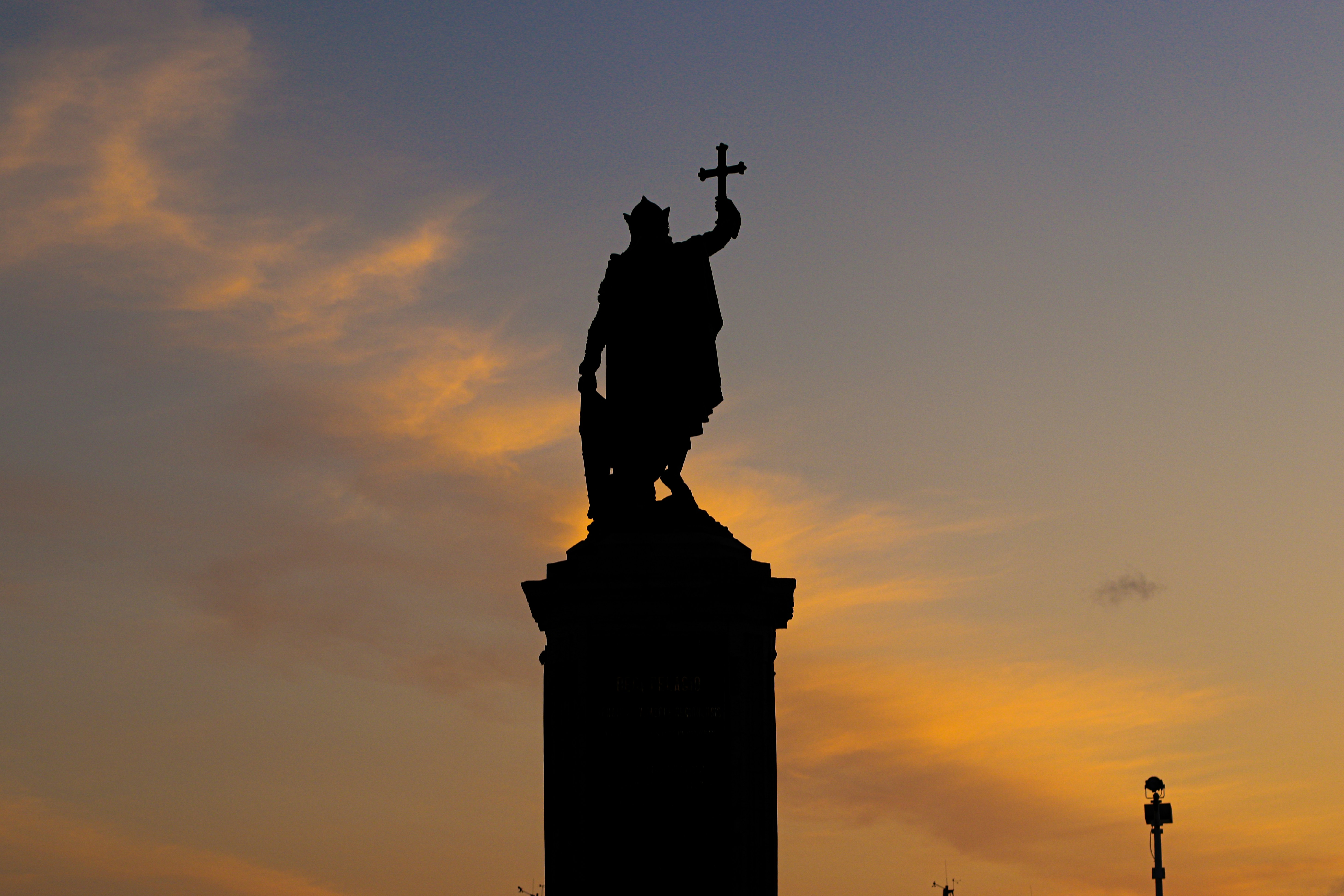 Statue of a man holding a cross on a building silhouetted against a vibrant sunset sky.