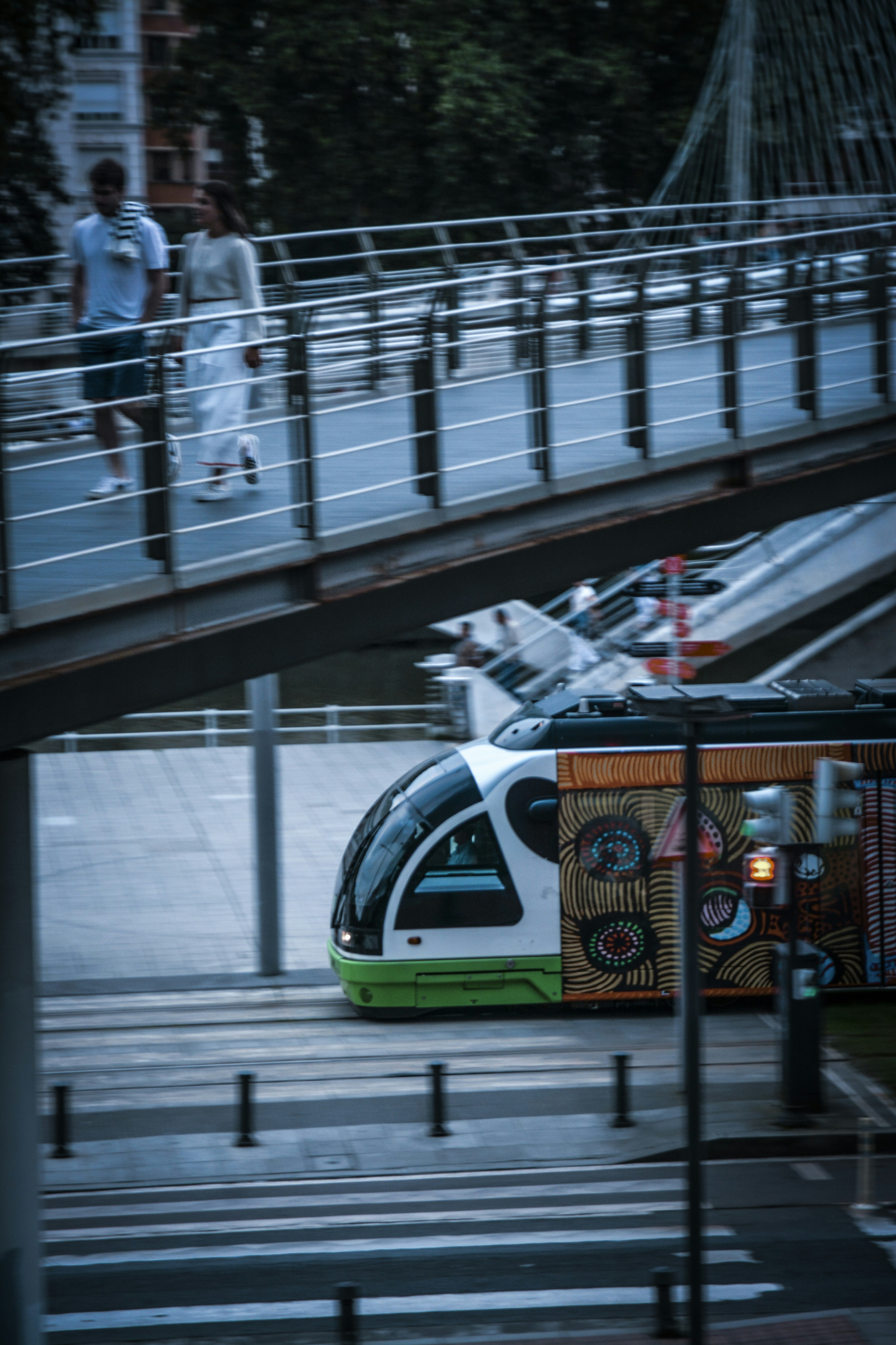 Modern tram passing beneath a pedestrian bridge in a cityscape.