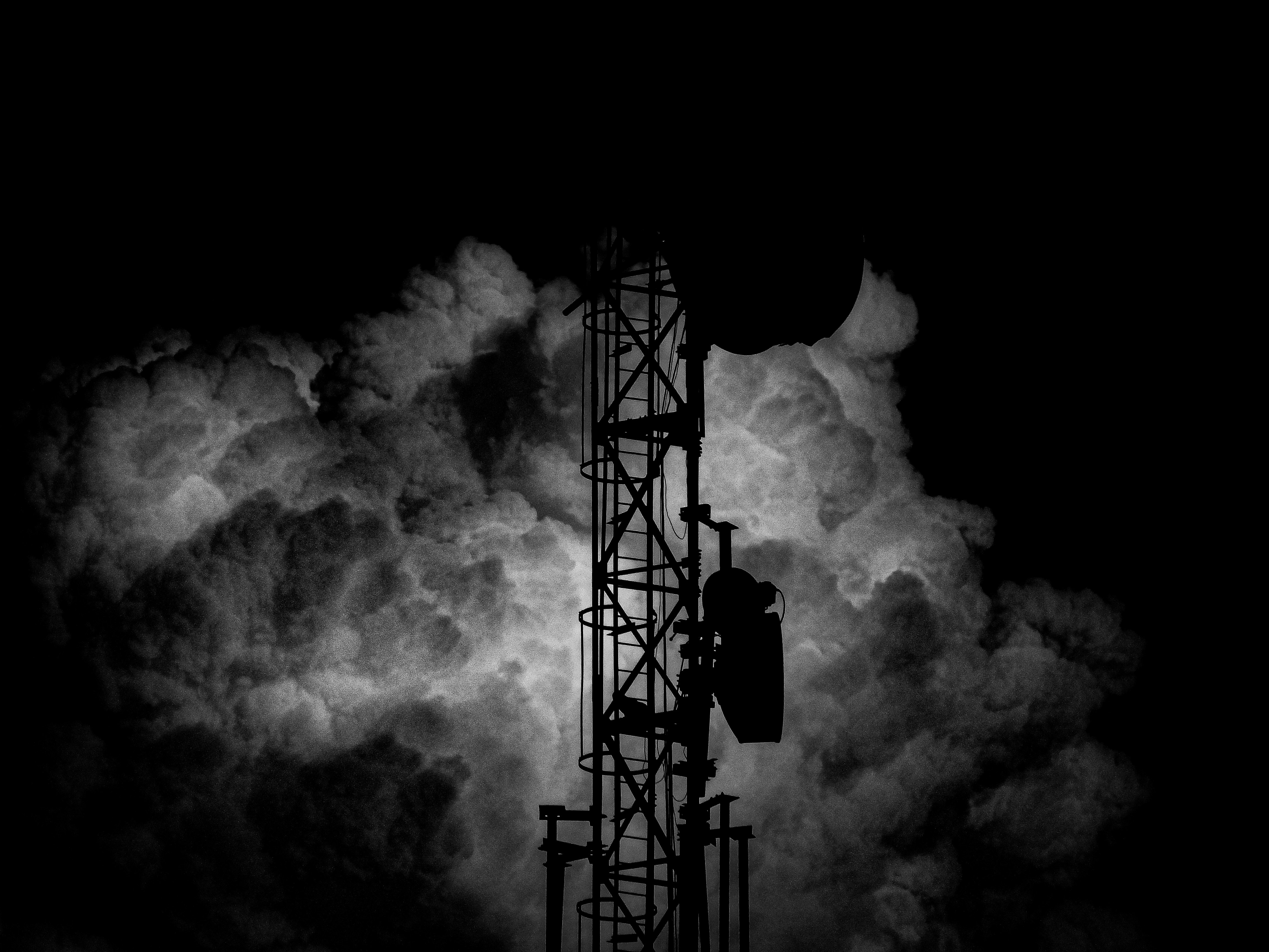 Telecommunication tower stands silhouetted against dramatic, swirling clouds in a monochrome palette.
