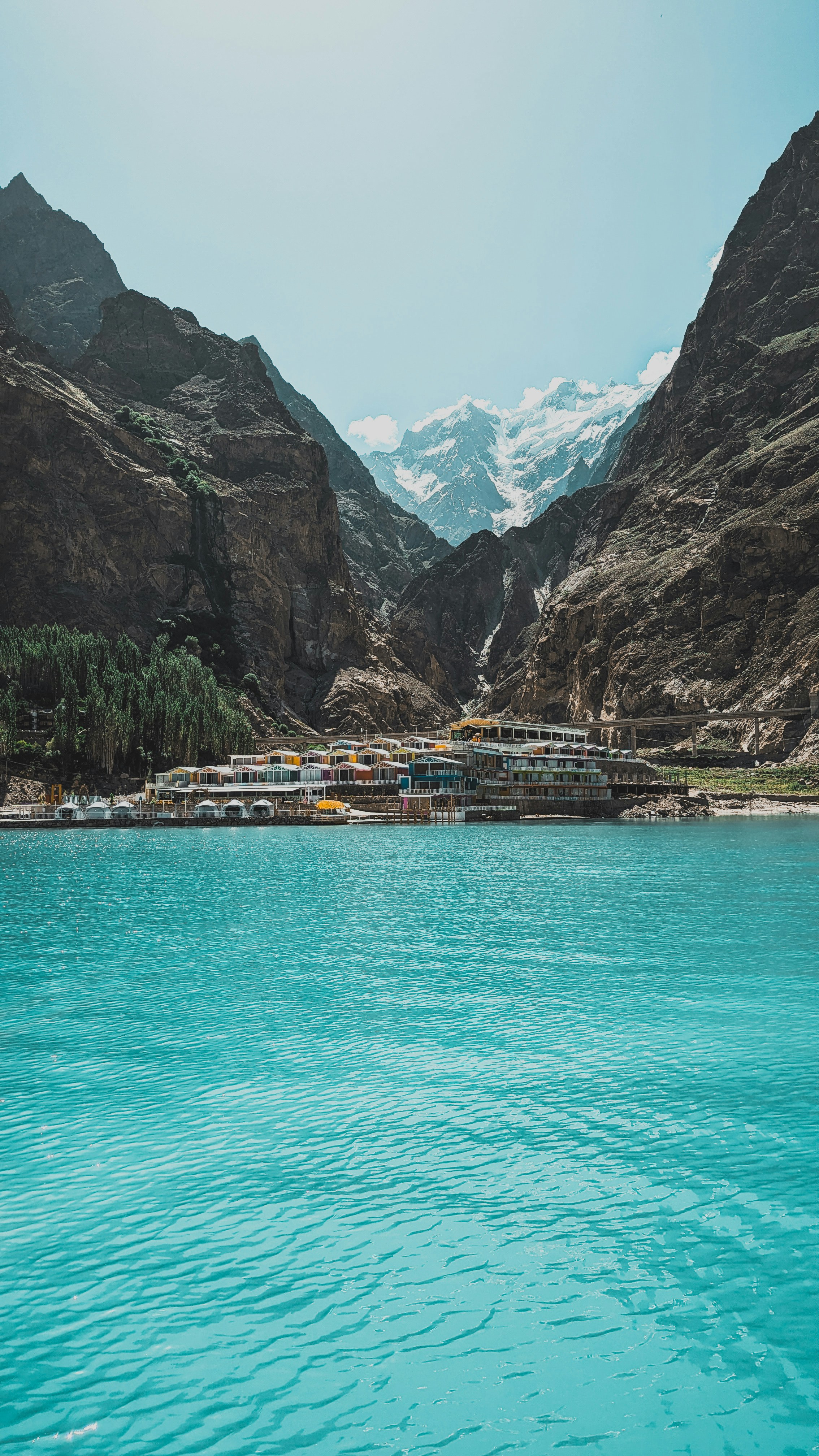 Turquoise lake with a stacked village at the waterline, set against dramatic rocky mountains and distant snow peaks.