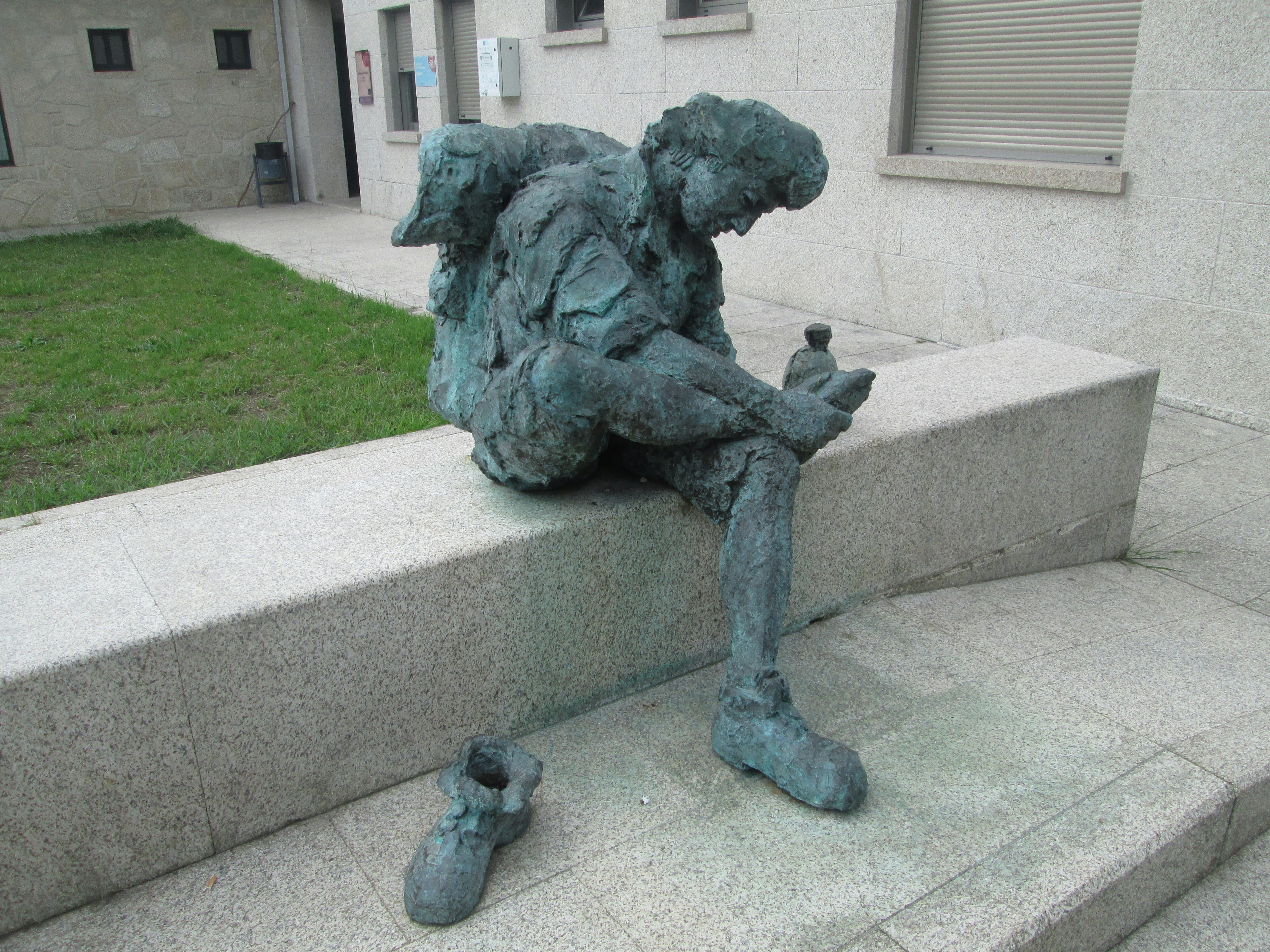 Blue-green patina sculpture of a seated figure with a backpack rests on a concrete ledge beside a building. A small shoe lies nearby on the ground.