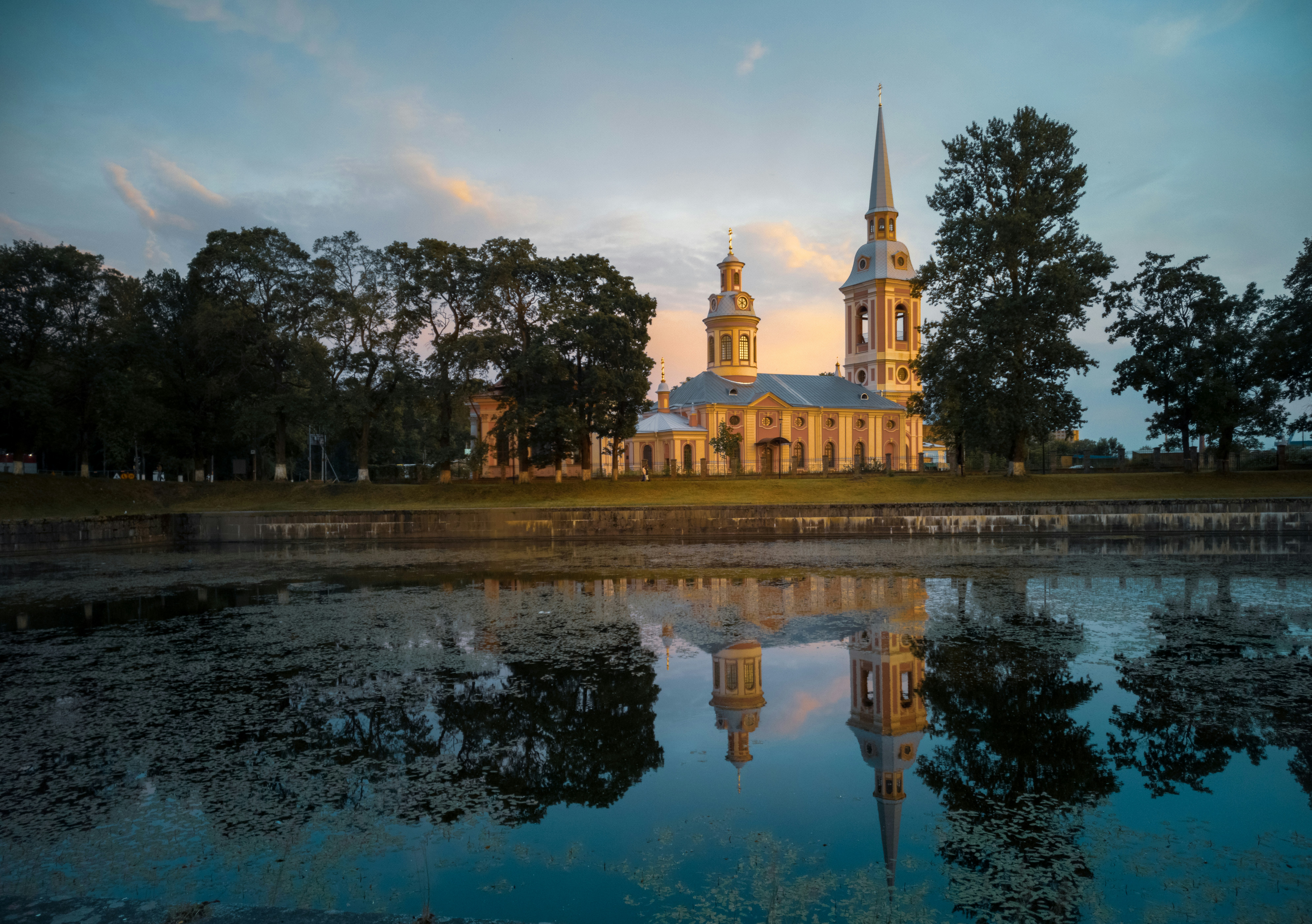 Cathedral with twin spires sits along a calm lakeside at dusk, bathed in warm light. Its reflection mirrors the façade in the still water.