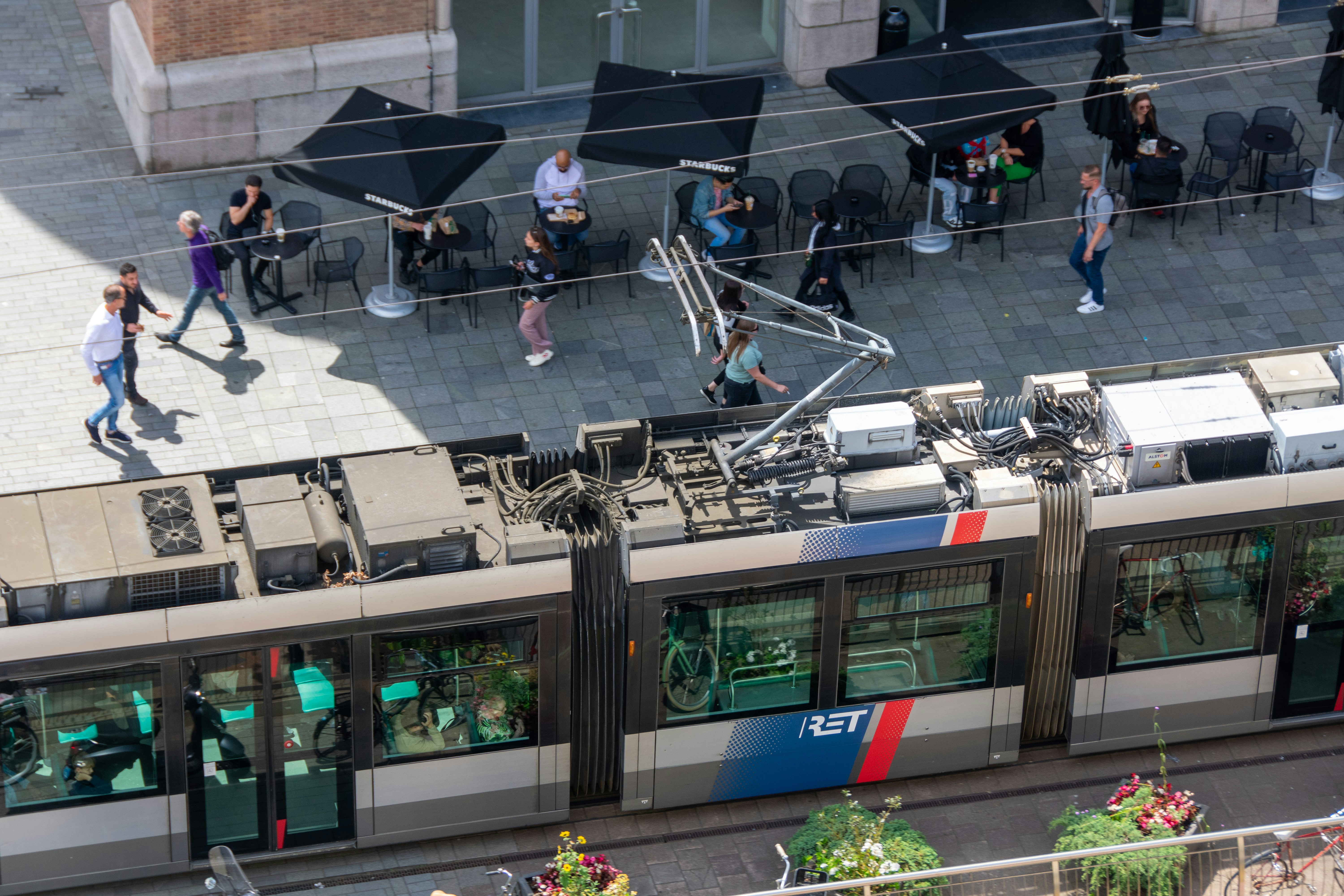 Overhead view of a tram navigating a city street with pedestrians walking nearby on a sunny day.