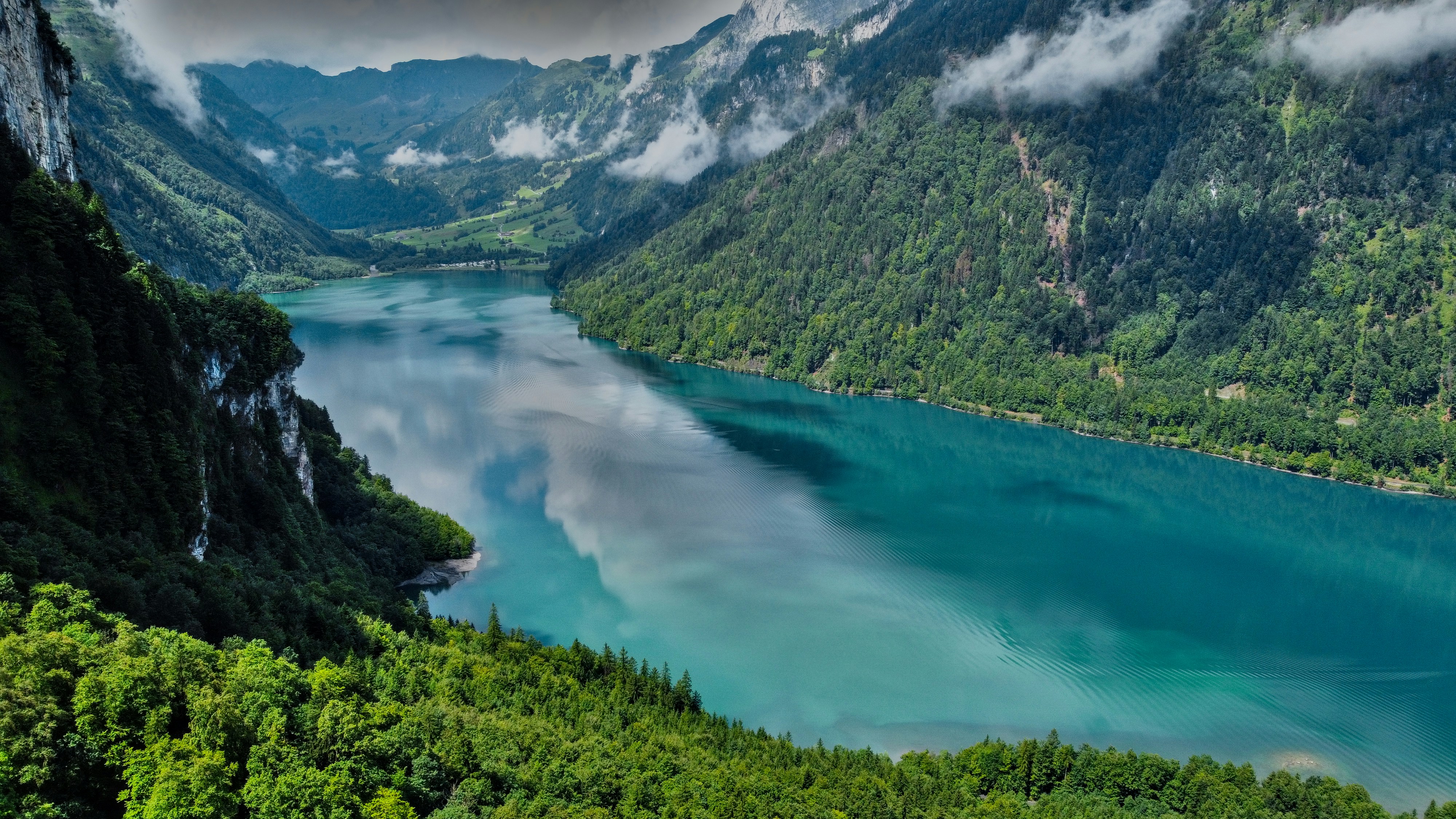 This breathtaking aerial image captures a tranquil alpine lake surrounded by lush, green mountains and shrouded in soft, low-hanging clouds. The vibrant turquoise water reflects the cloudy sky, creating a mesmerizing blend of colors that contrast with the deep greens of the forested slopes. The composition, with its sweeping view of the landscape, exudes a sense of peace and natural beauty, making it a striking visual masterpiece.