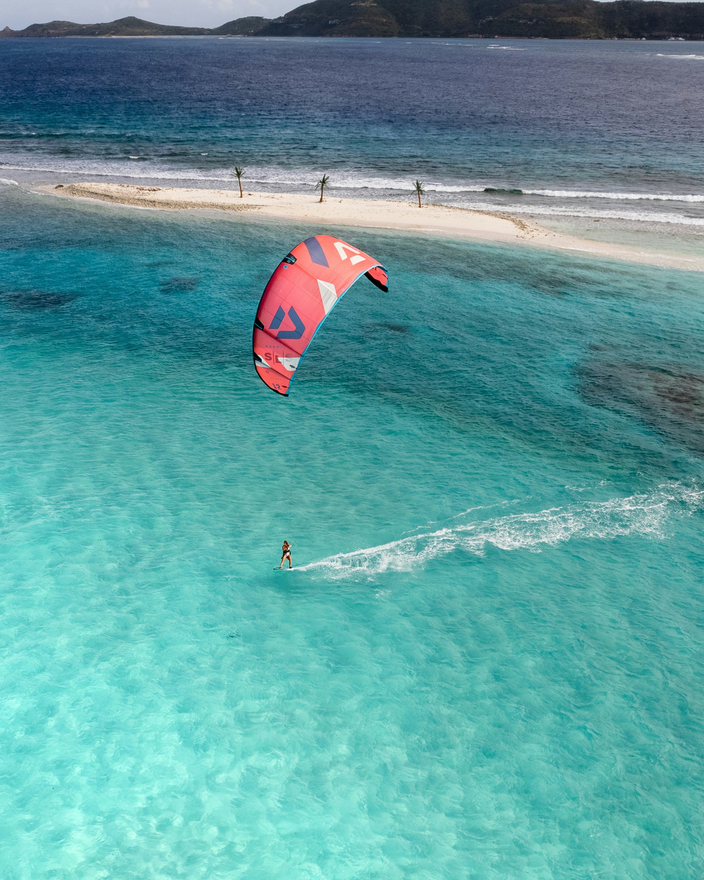 A kiteboarder glides across crystal-clear turquoise waters, with a vibrant red kite soaring above. The serene beach and gentle waves create a perfect backdrop for this exhilarating sport.