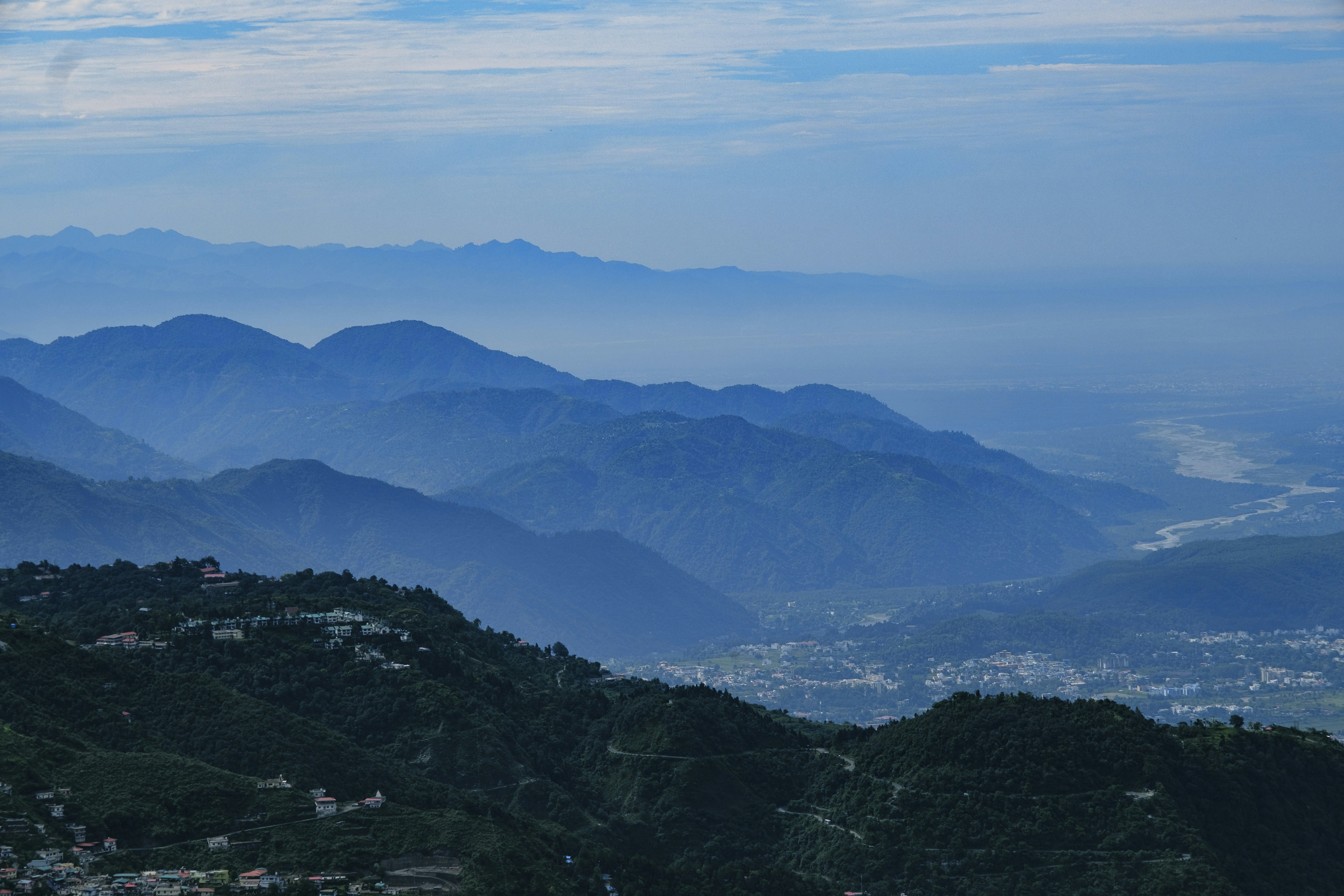 Layered mountain ranges shrouded in mist under a soft blue sky, revealing a serene valley below.