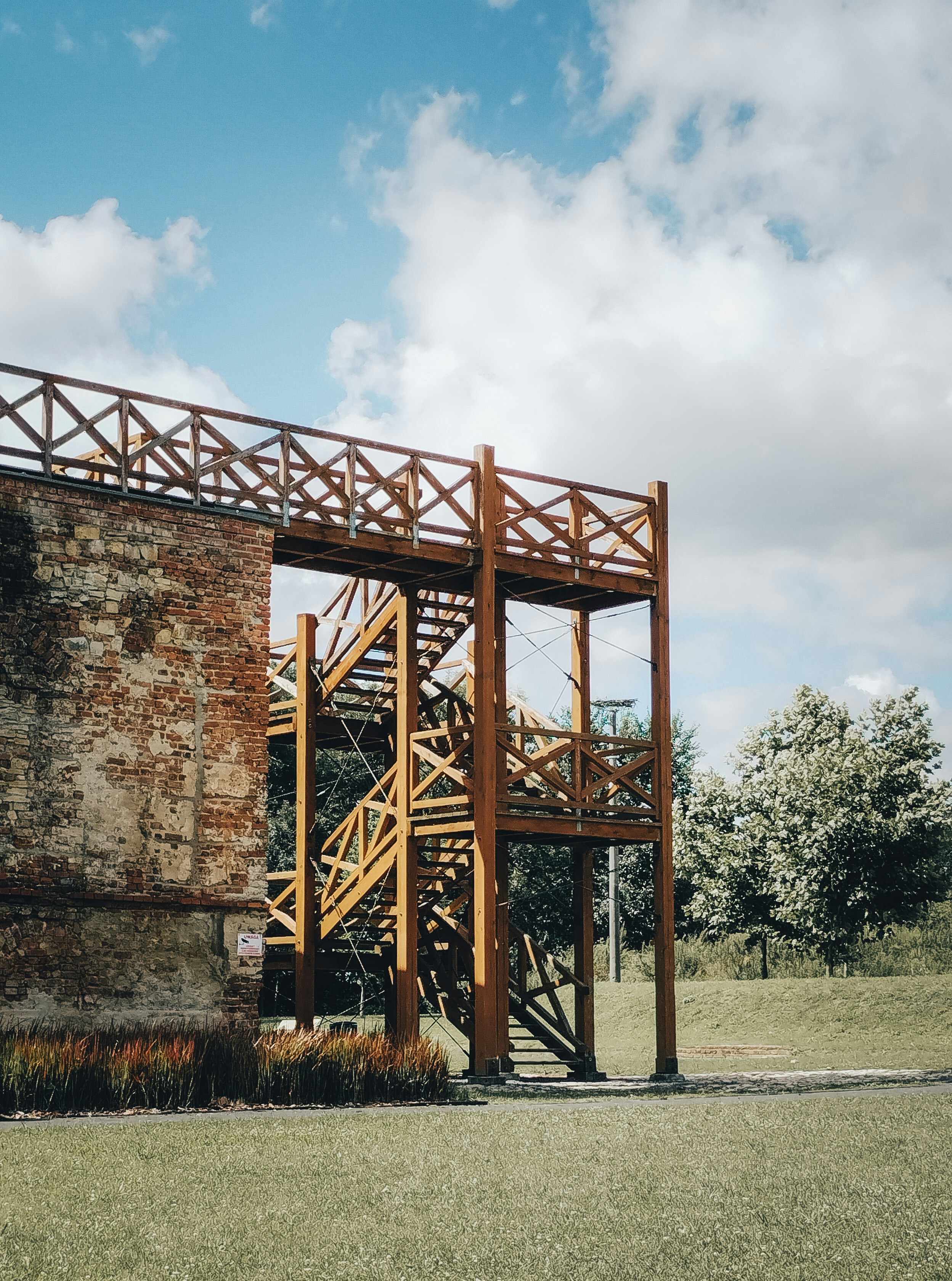 A wooden staircase framework attached to a brick wall sits on a grassy field beneath a bright blue sky.