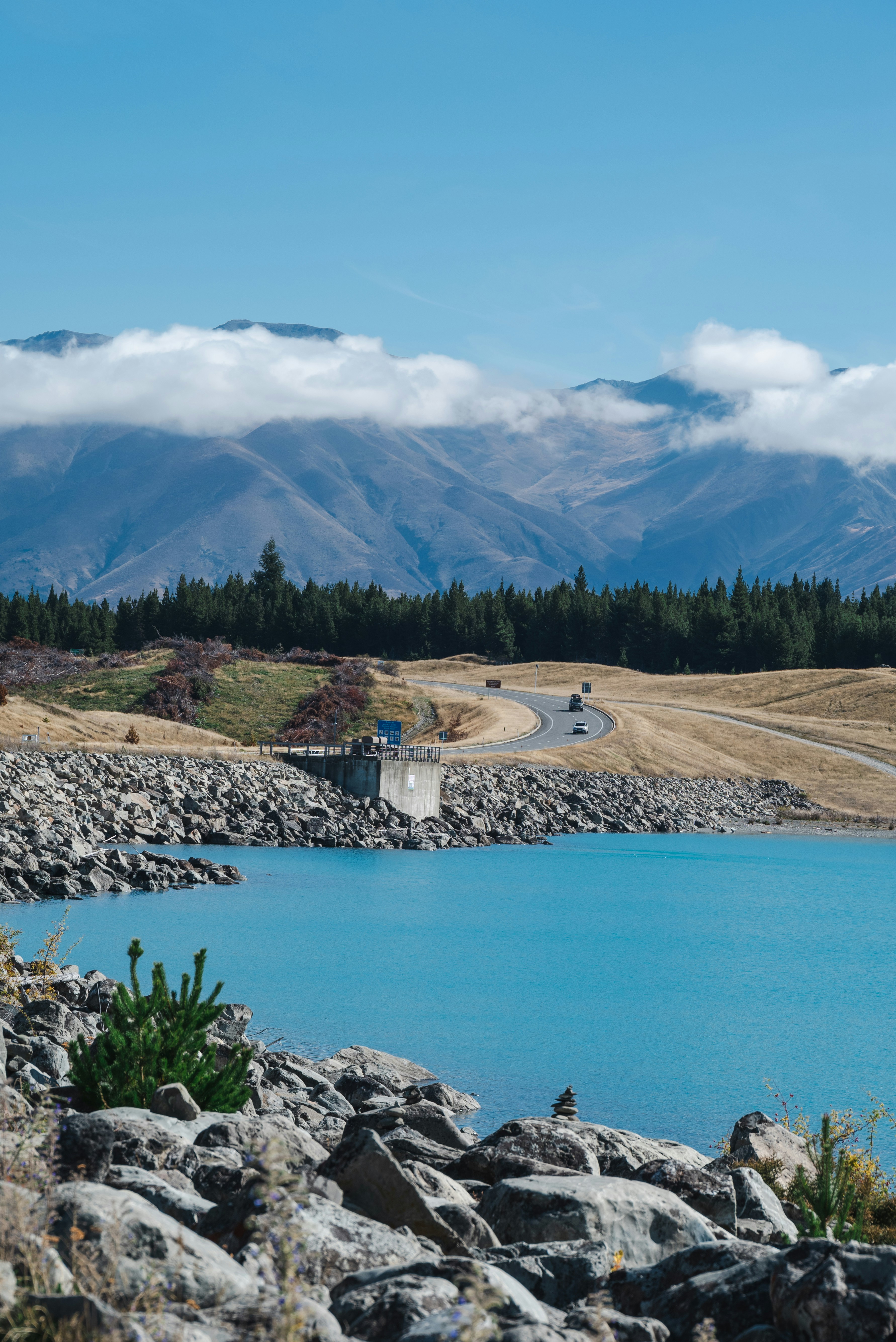 A winding road curves along a tranquil lake, framed by rocky shores and distant mountains under a clear blue sky.
