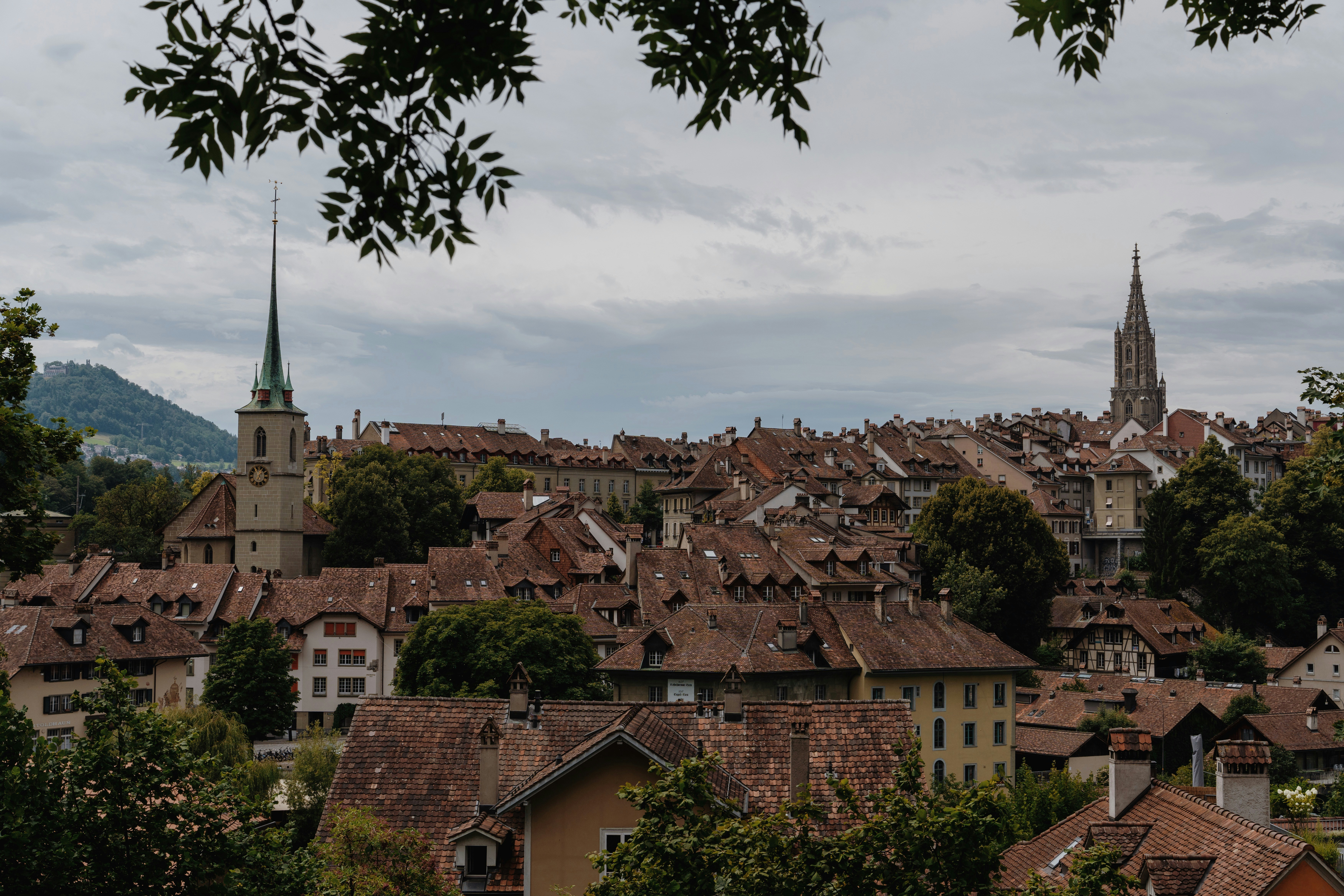 Panoramic view of a historic town with clustered red-tiled rooftops and two prominent church spires under a cloudy sky.