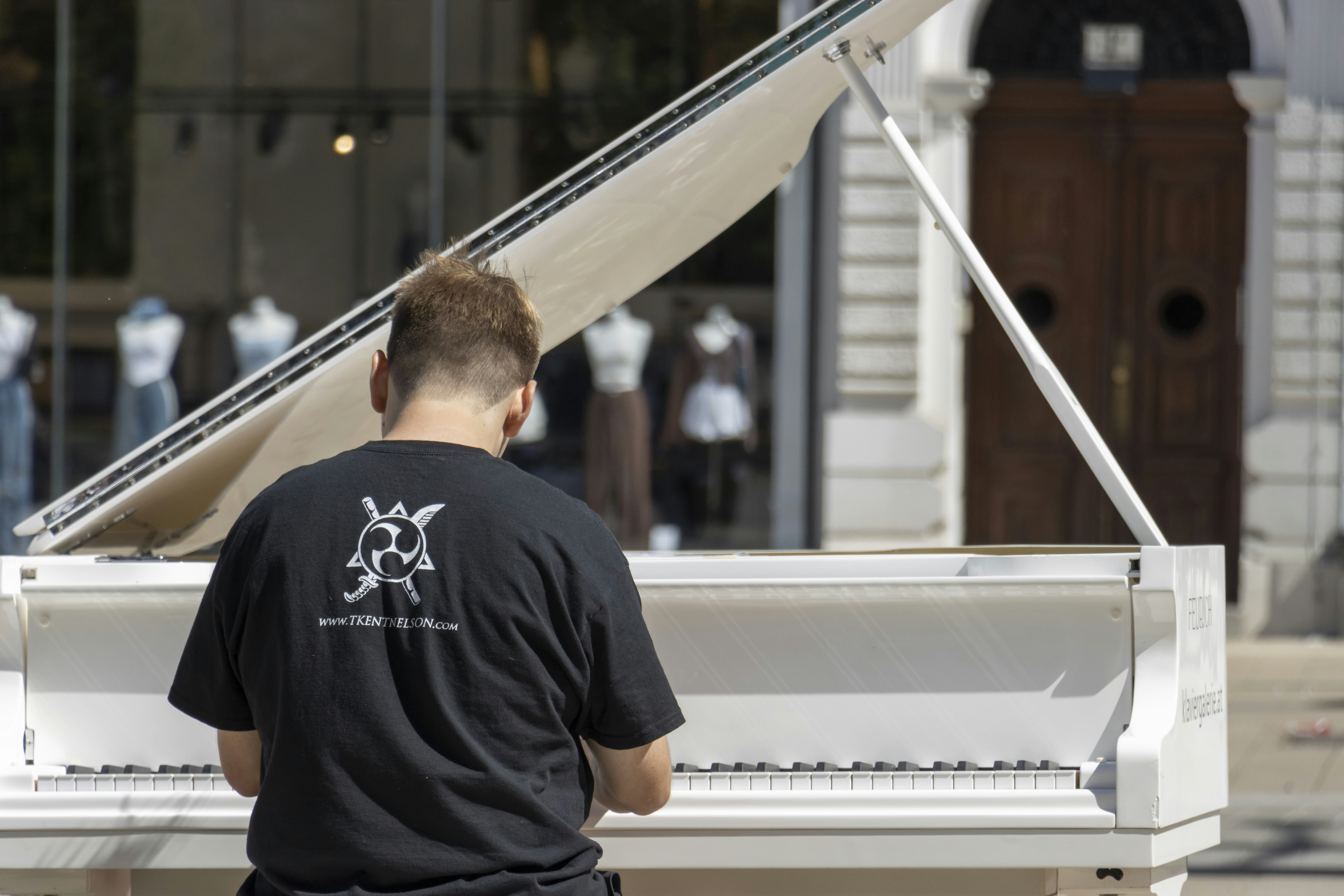 Midway in a public plaza, a pianist tunes the air beside a white grand