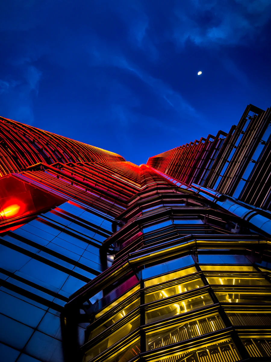 Low-angle view of a modern high-rise glowing red, blue, and yellow against a deep twilight sky, with a small moon overhead.