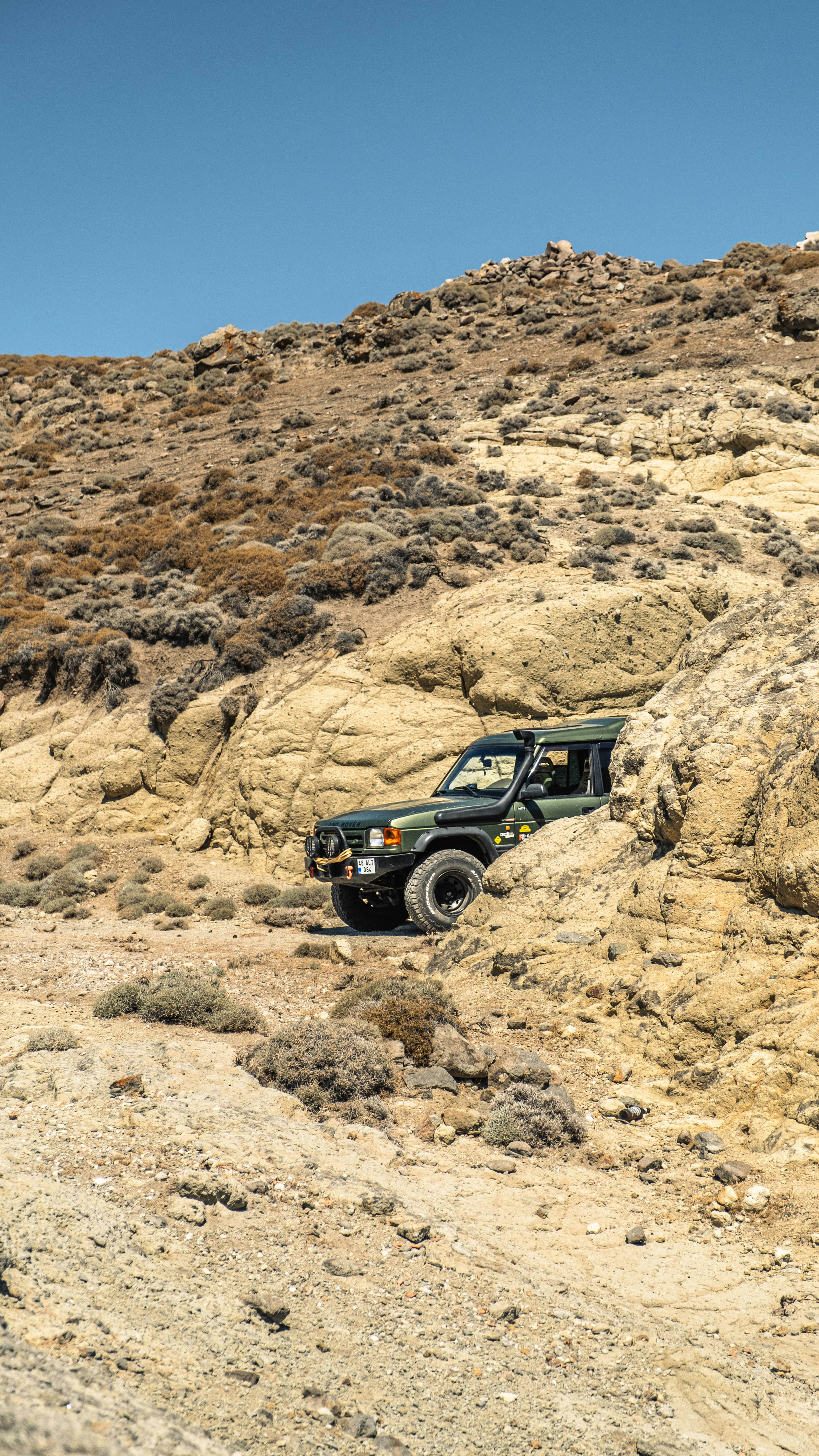 A rugged off-road vehicle navigating through rocky terrain under a clear blue sky. The landscape showcases earthy tones and textures, emphasizing the adventure spirit.