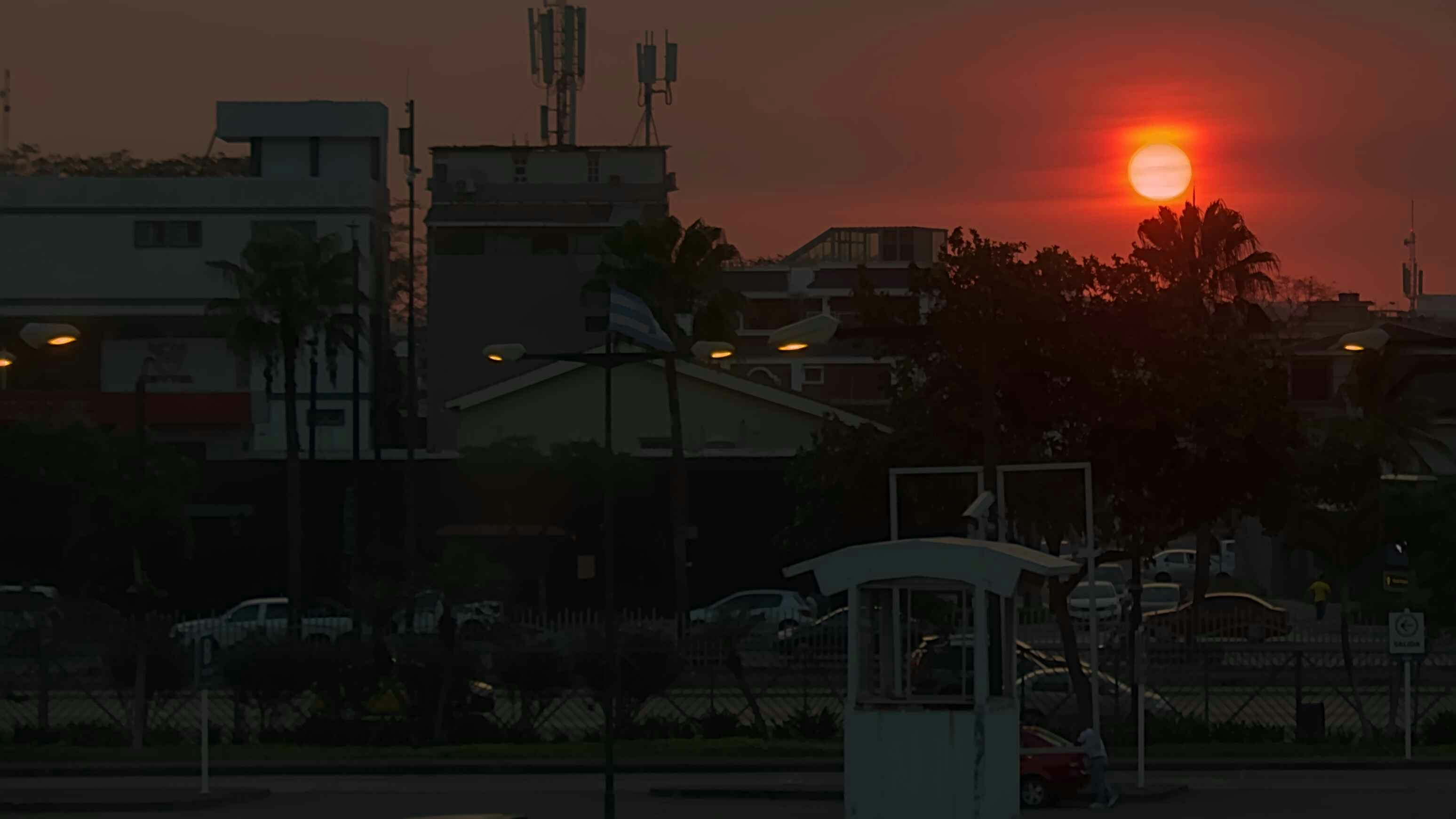 Sunset casts a vibrant red hue over a townscape with silhouetted buildings and trees.
