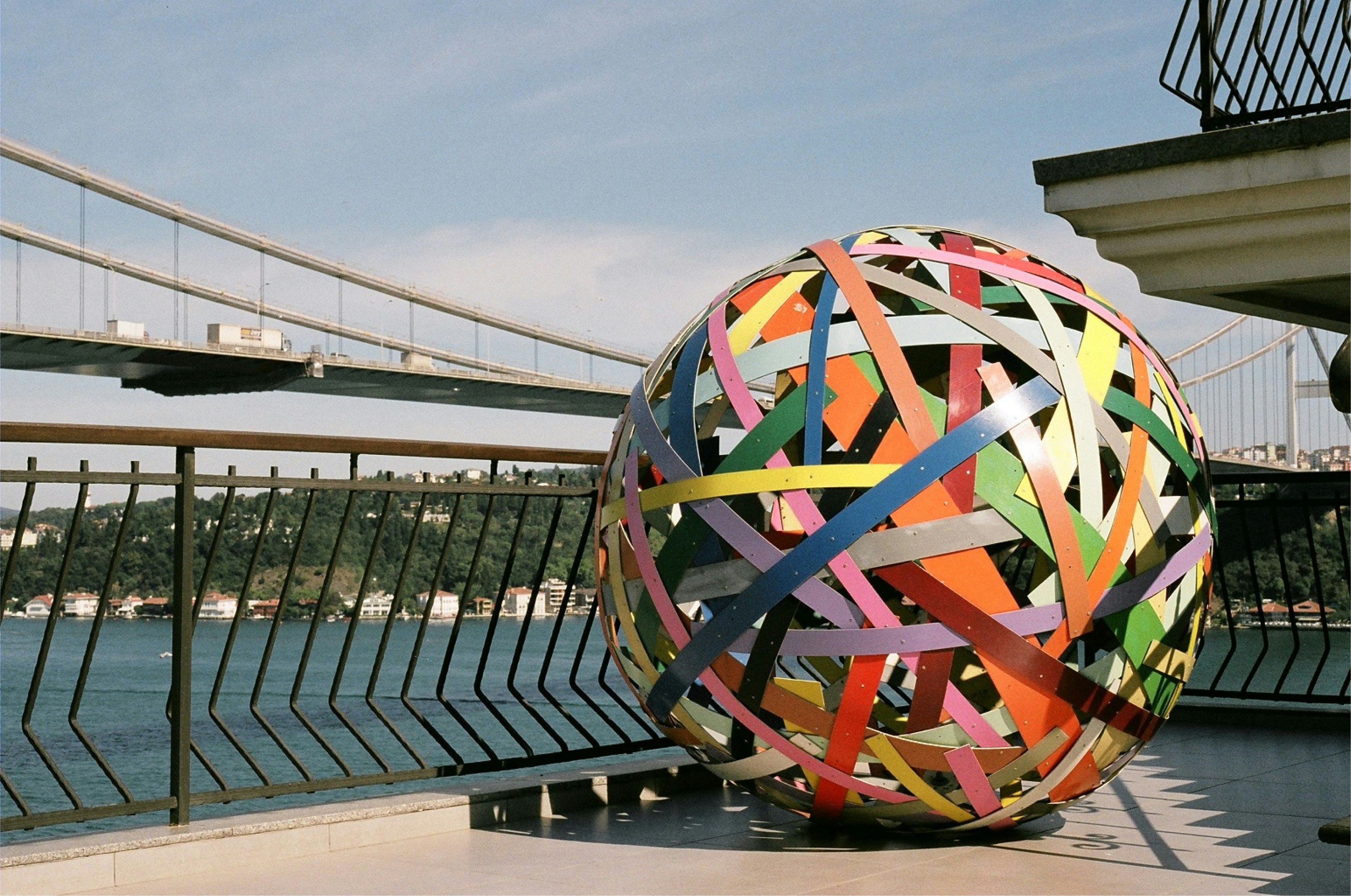 Colorful spherical sculpture composed of interwoven ribbons on a riverside deck with a suspension bridge in the background.