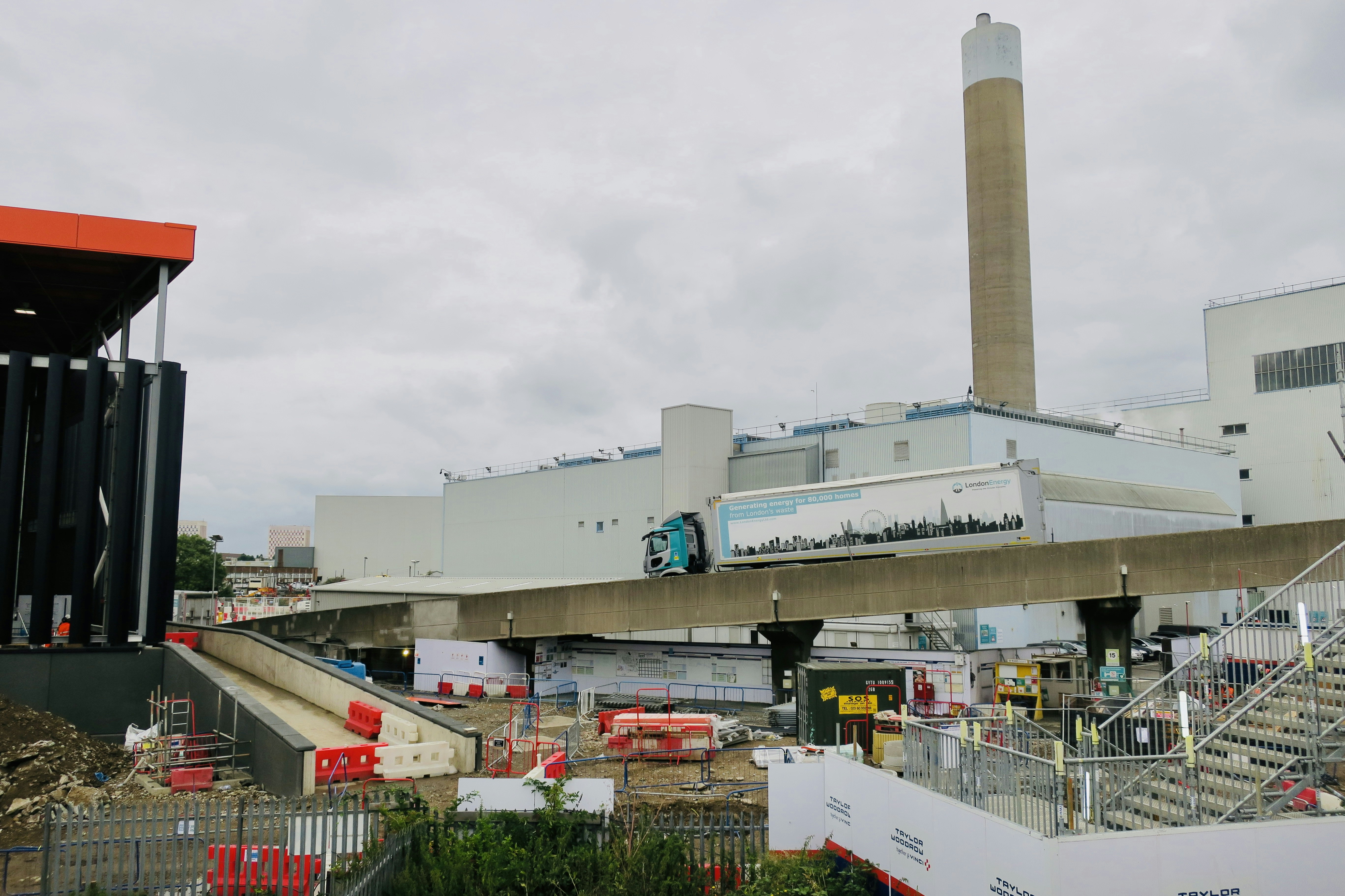 Construction site under a concrete overpass with machinery, fencing, and a distant industrial building.