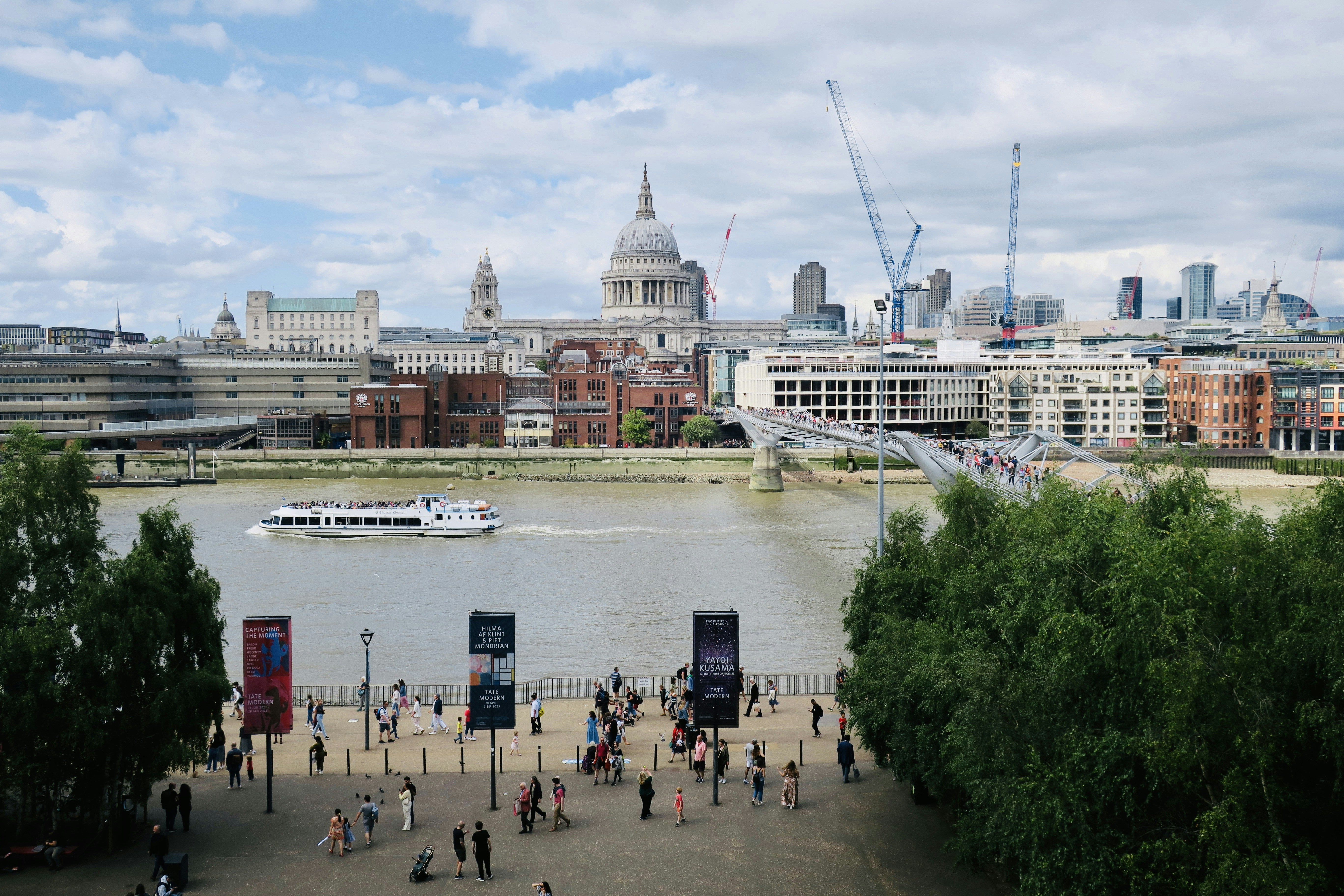 Bustling riverfront with pedestrians along the South Bank, boat cruising the Thames, and the city skyline including St. Paul’s dome in the distance.