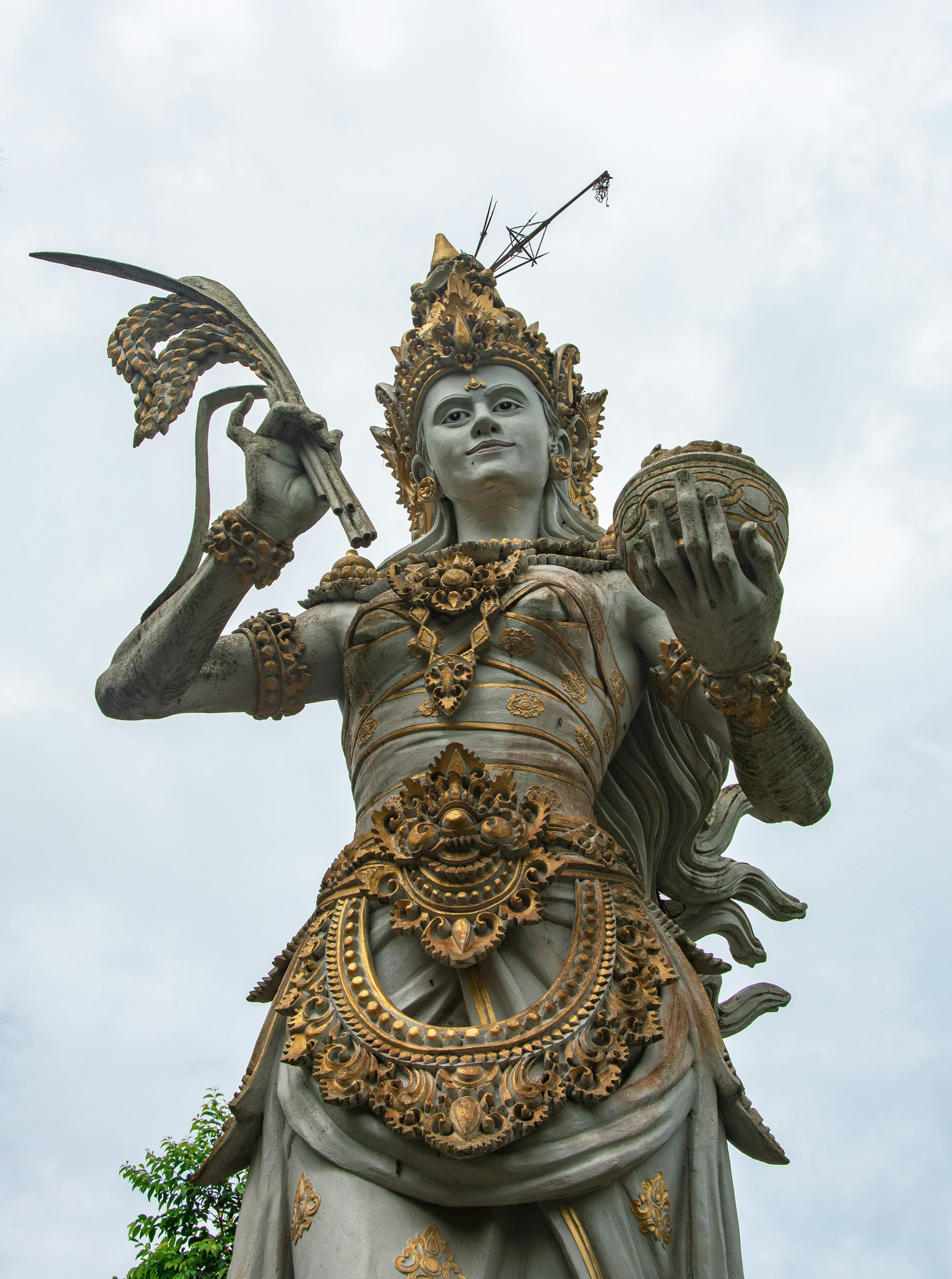 Gilded Sentinel: A Stone Deity with Curved Blade and a Bowl Under a Clouded Sky