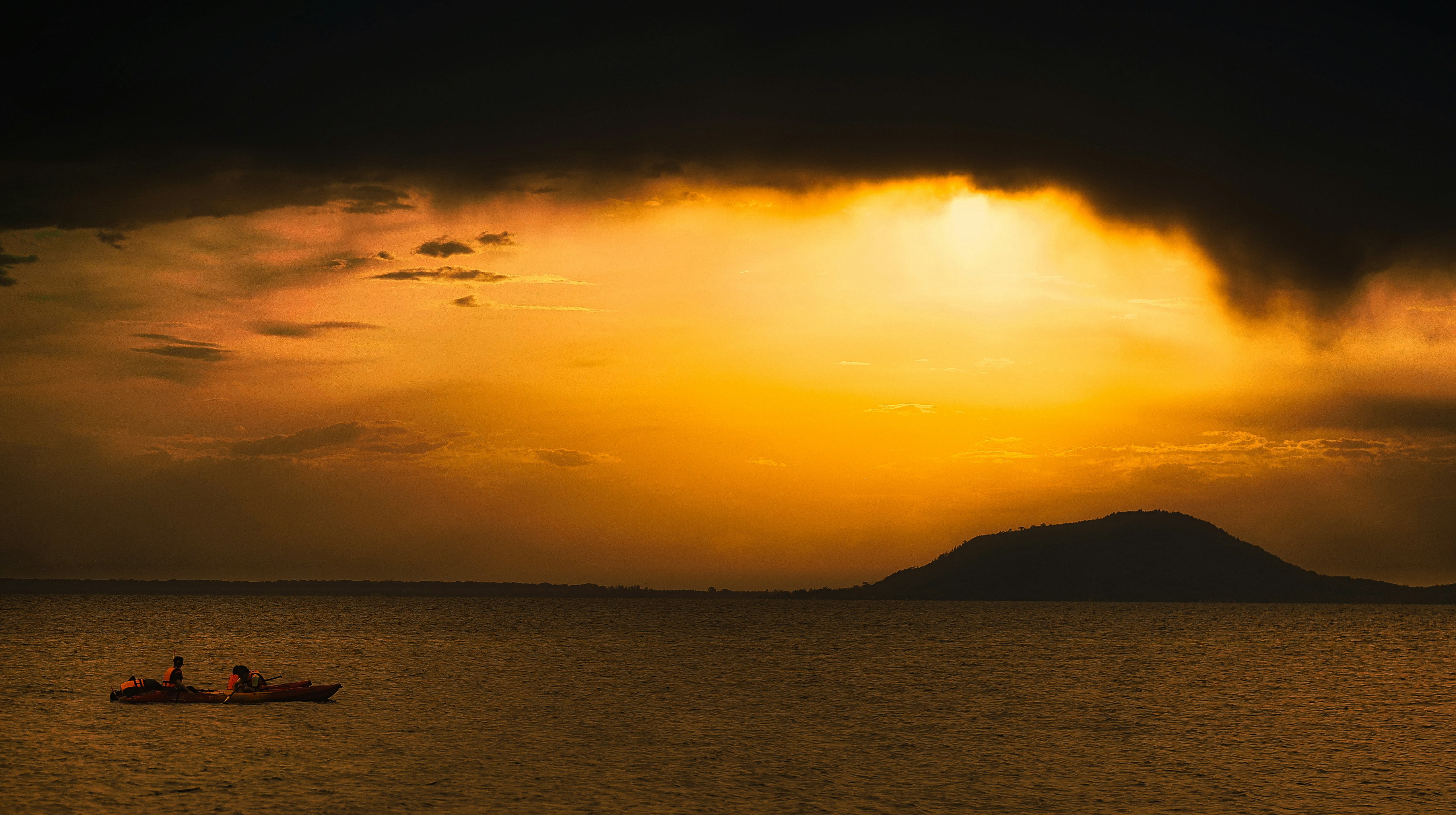 Two kayakers navigate tranquil waters under a dramatic sunset, with a silhouette of a distant island. The sky is painted in warm hues, contrasting with the dark clouds above.