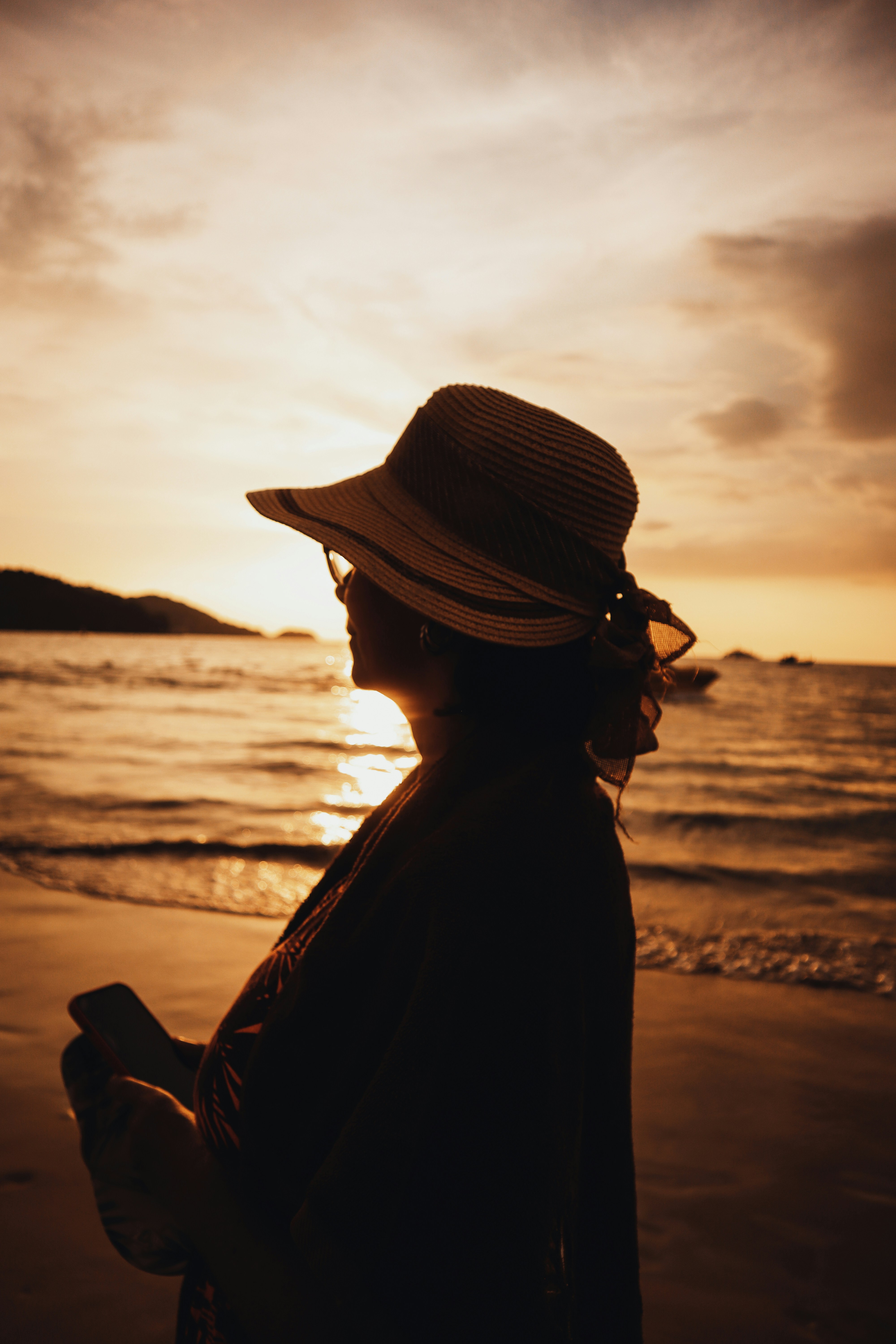 Silhouette of a woman in a sun hat standing on a beach at sunset, with gentle waves in the background.