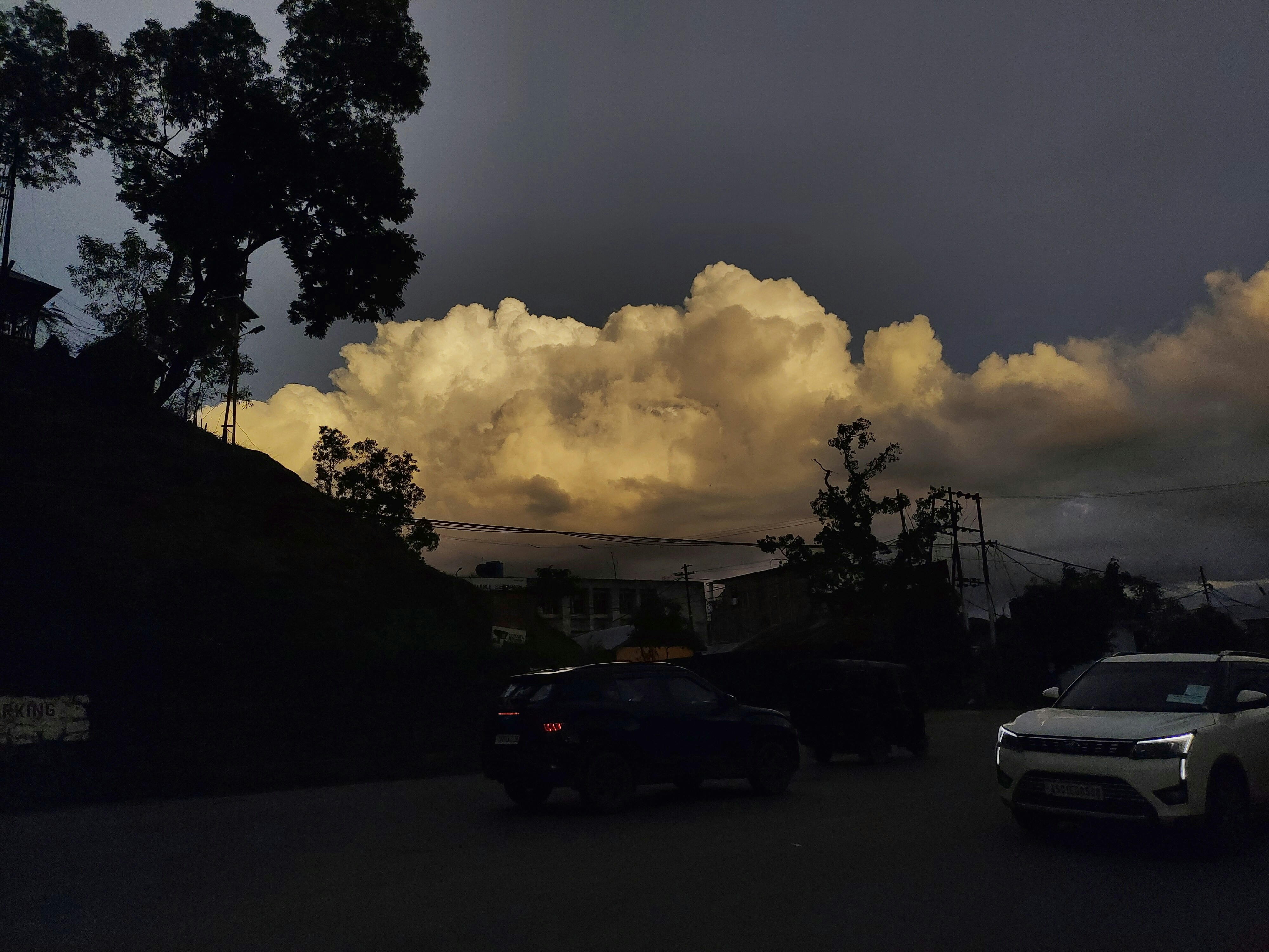 Dusk photograph of a dim parking lot with a row of cars beneath dramatic gold-tinged clouds. The scene emphasizes contrast between the dark foreground and the luminous sky.