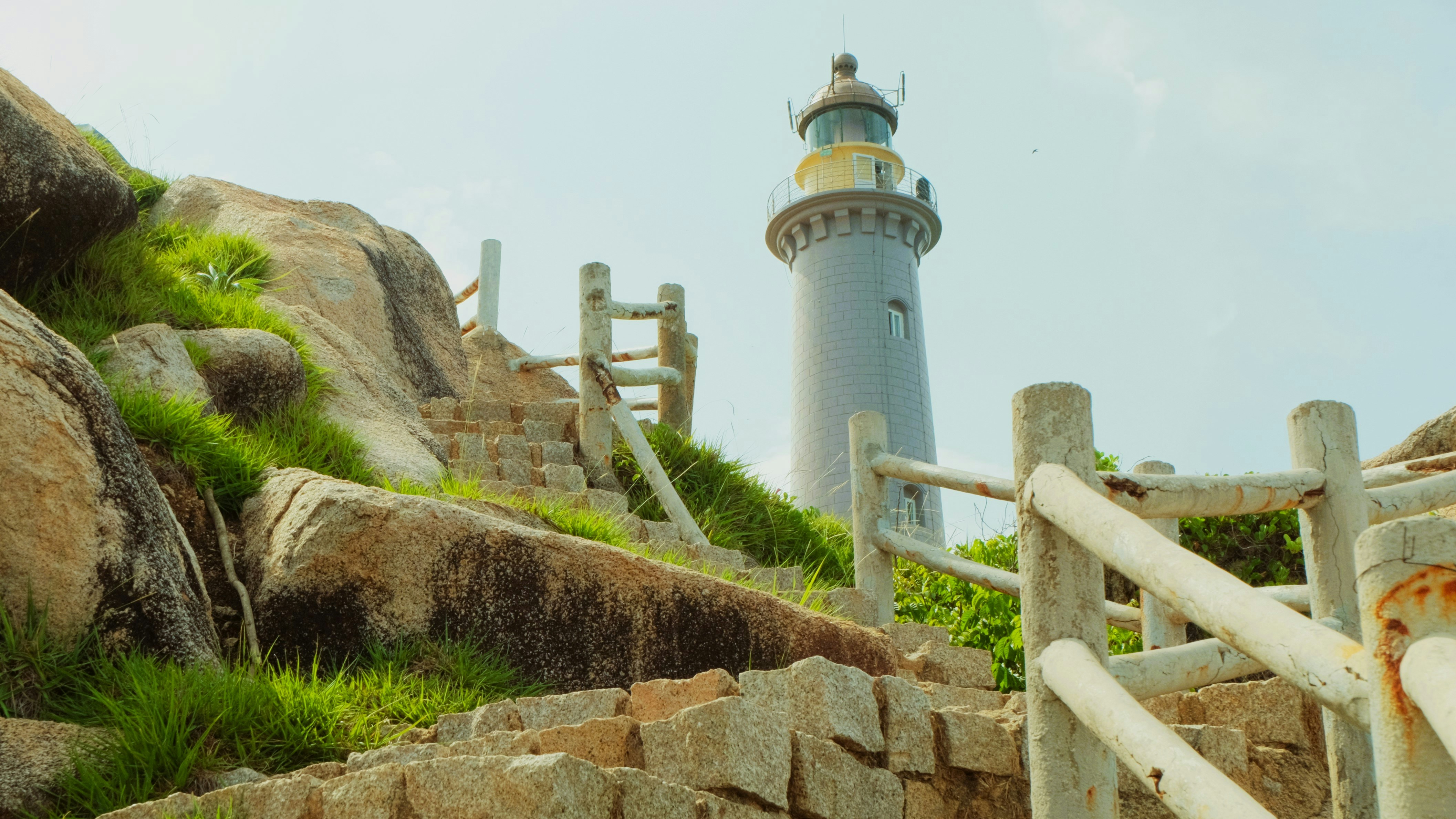 Lighthouse standing tall amidst rocky terrain and lush greenery, with a winding staircase leading up to it.