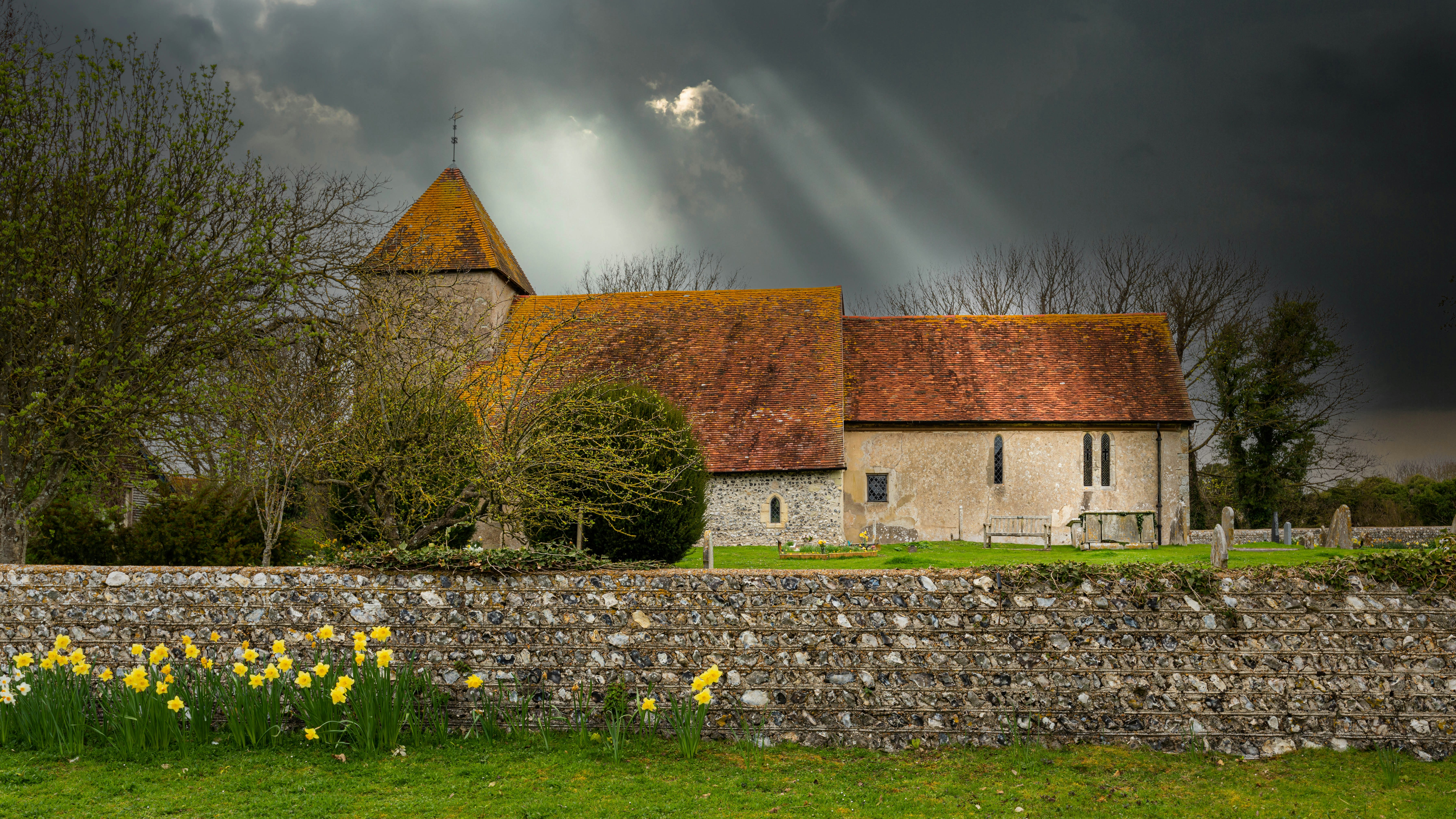 Sunbeams break through dark clouds, illuminating an ancient church and surrounding foliage.