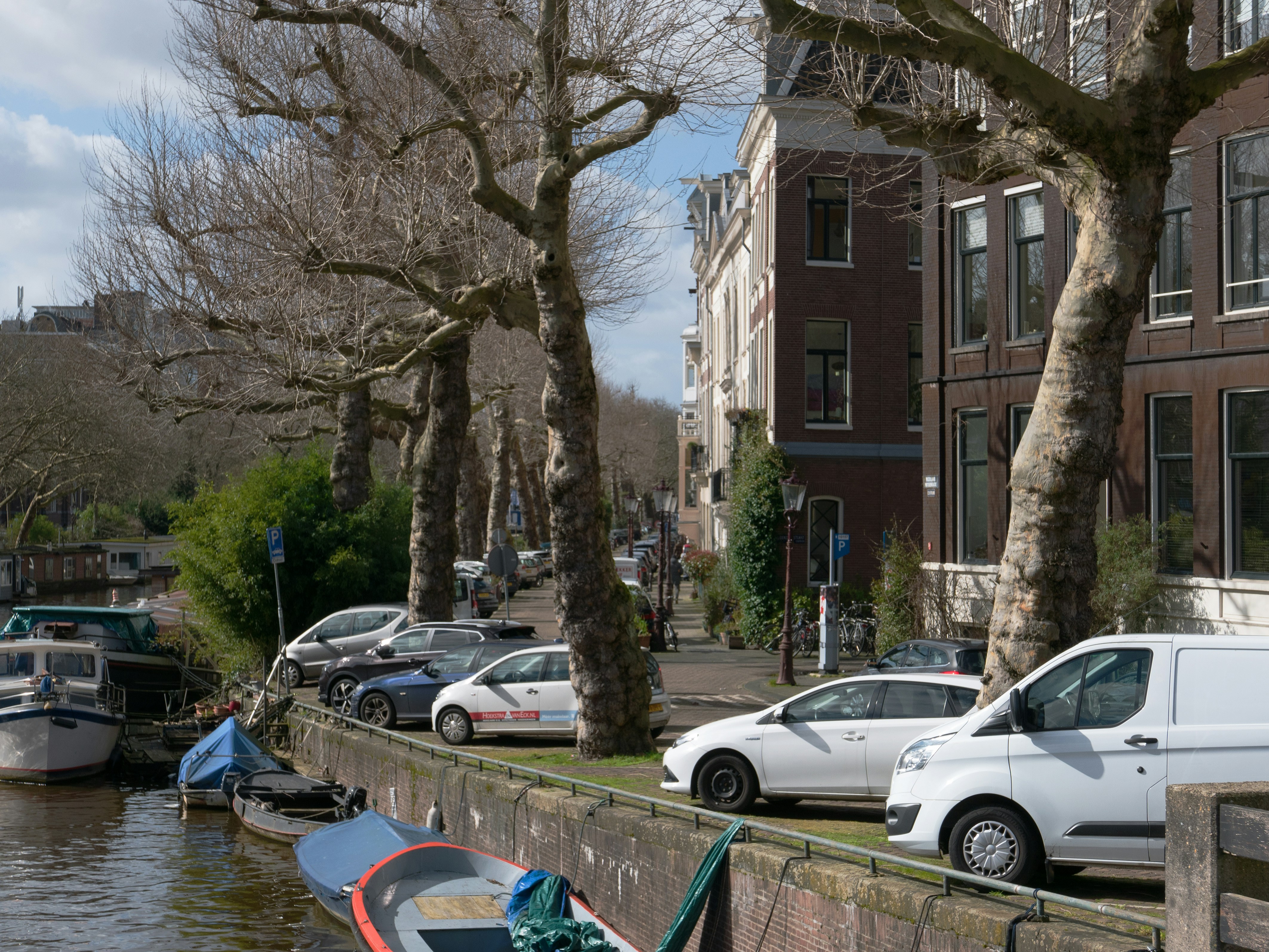 A tranquil canal scene featuring lined trees and parked vehicles along the waterfront, showcasing everyday life in a city environment.