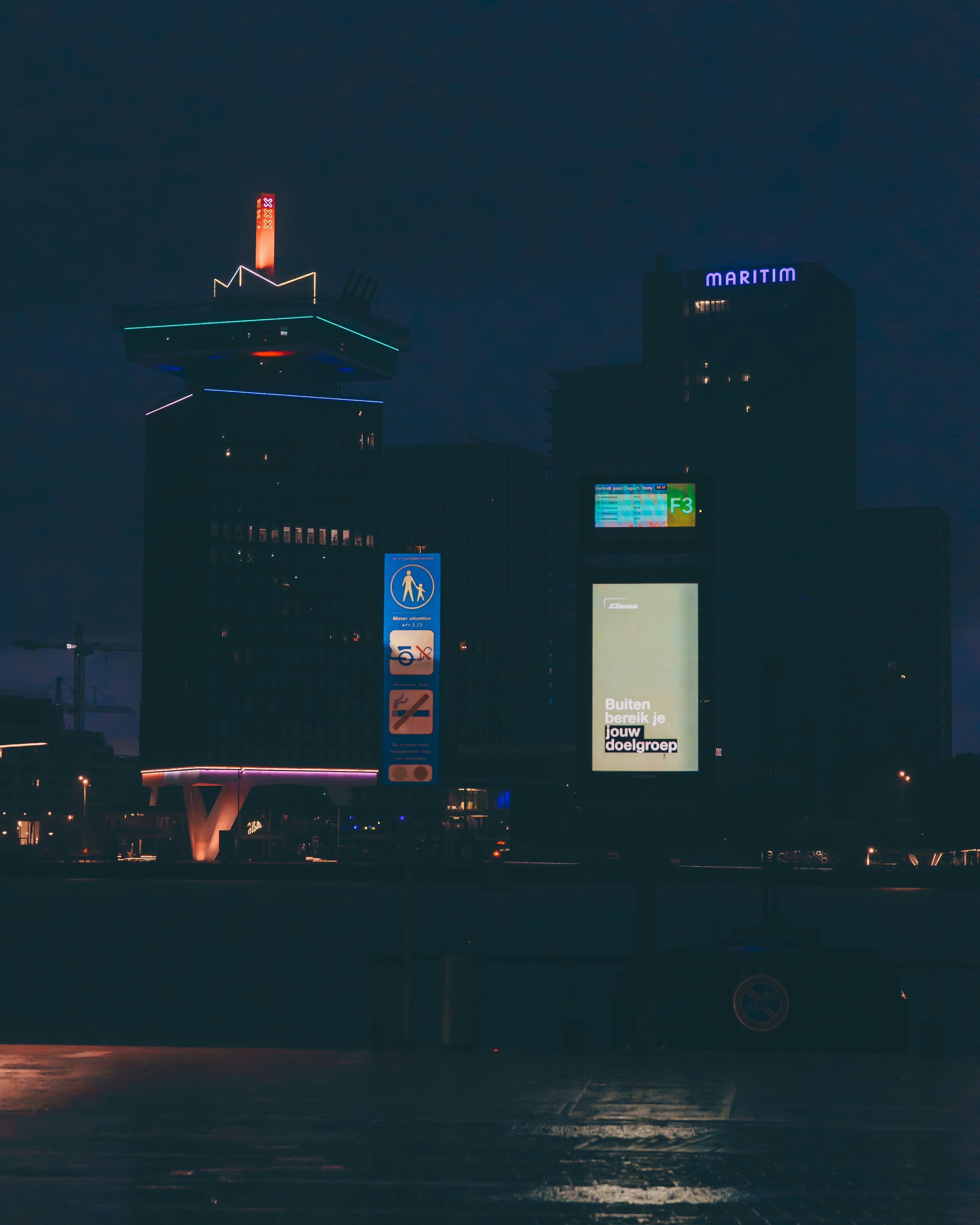 Neon-lit buildings against a dark evening sky with reflections on wet pavement.
