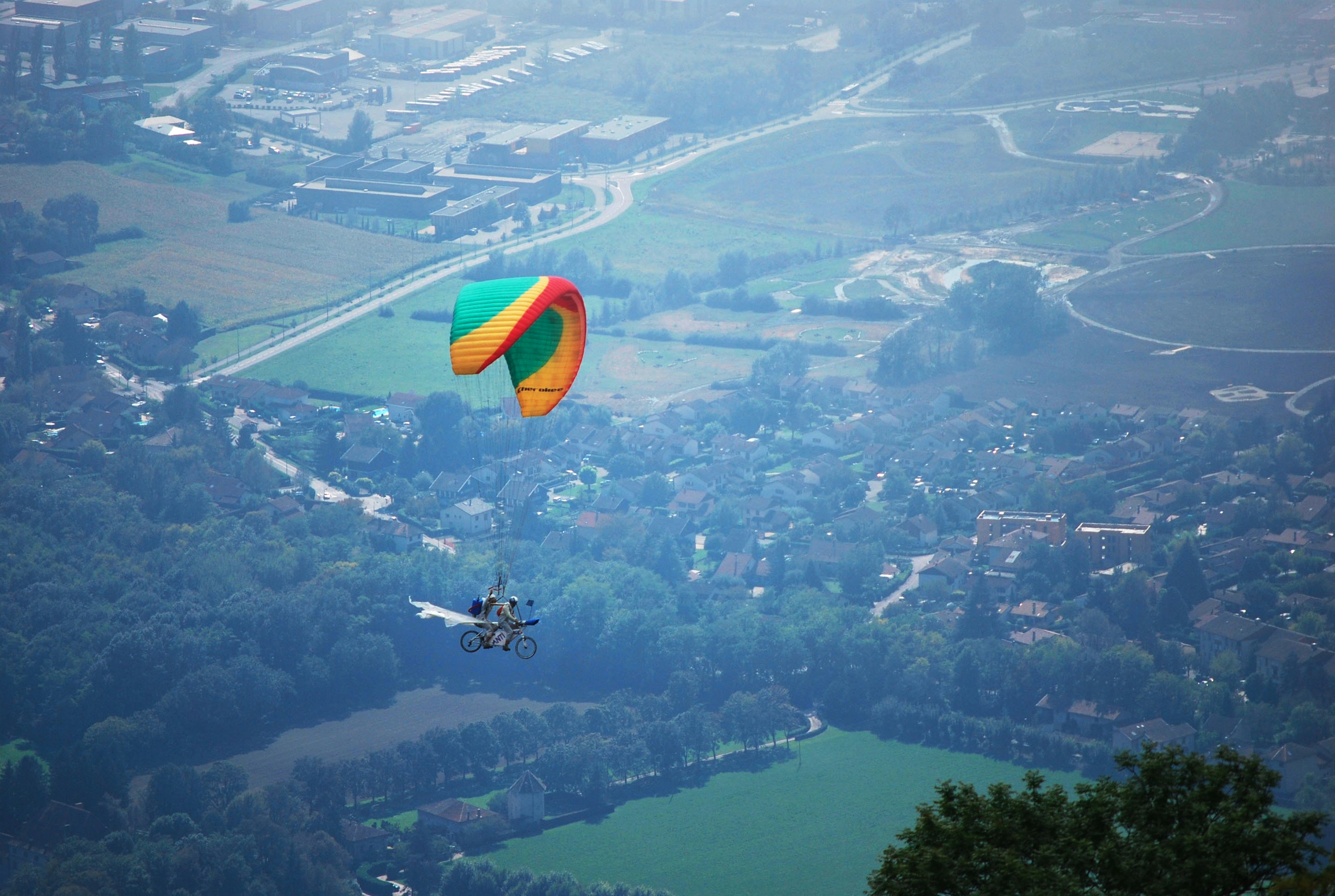 Aerial photograph of a rainbow paraglider gliding over a patchwork valley dotted with fields and a small town.