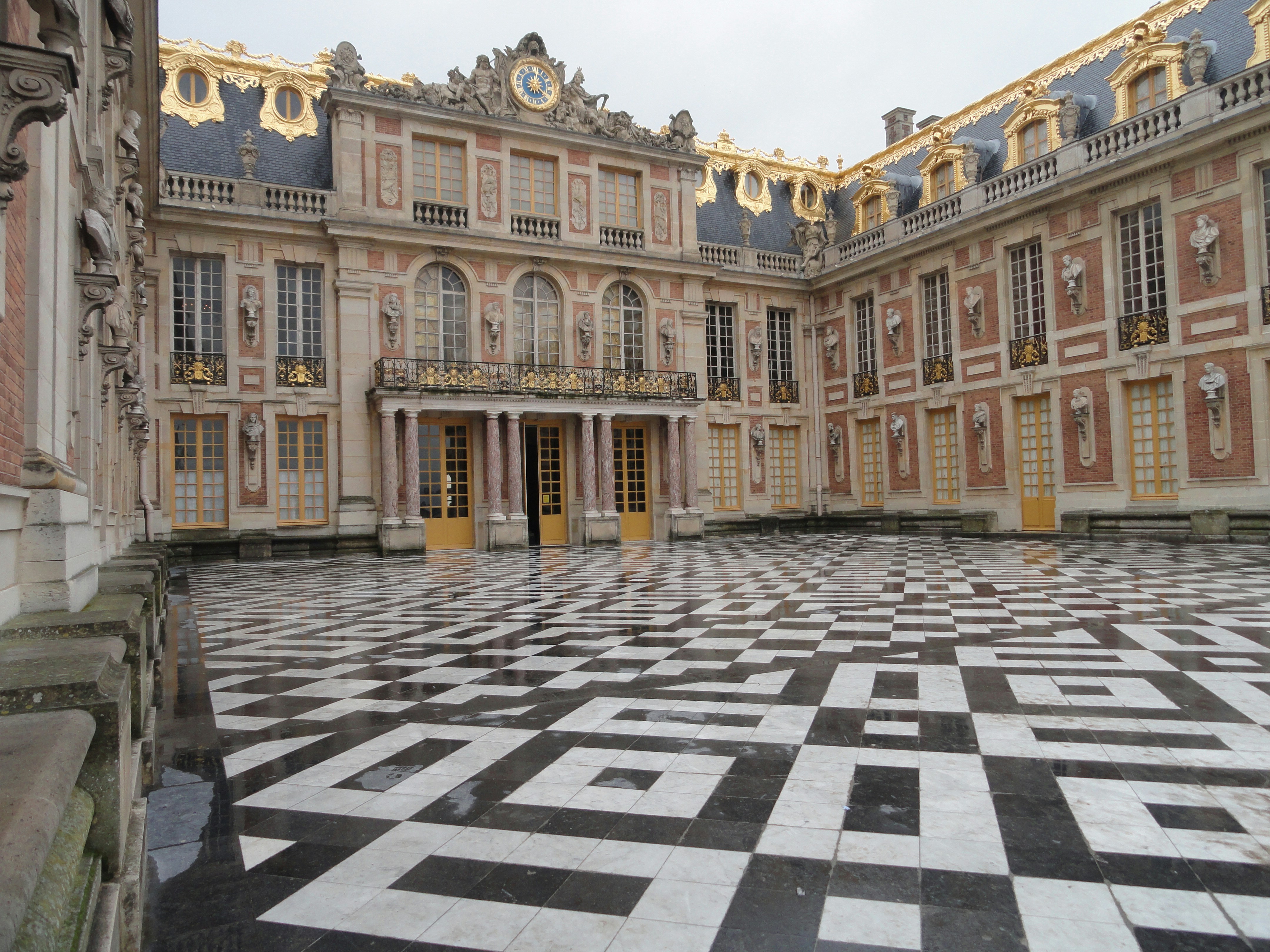 Palatial courtyard dominated by a bold black-and-white checkerboard floor that leads the eye toward ornate façades and gilded doors. Bright daylight highlights the symmetry and decorative details of the Baroque architecture.