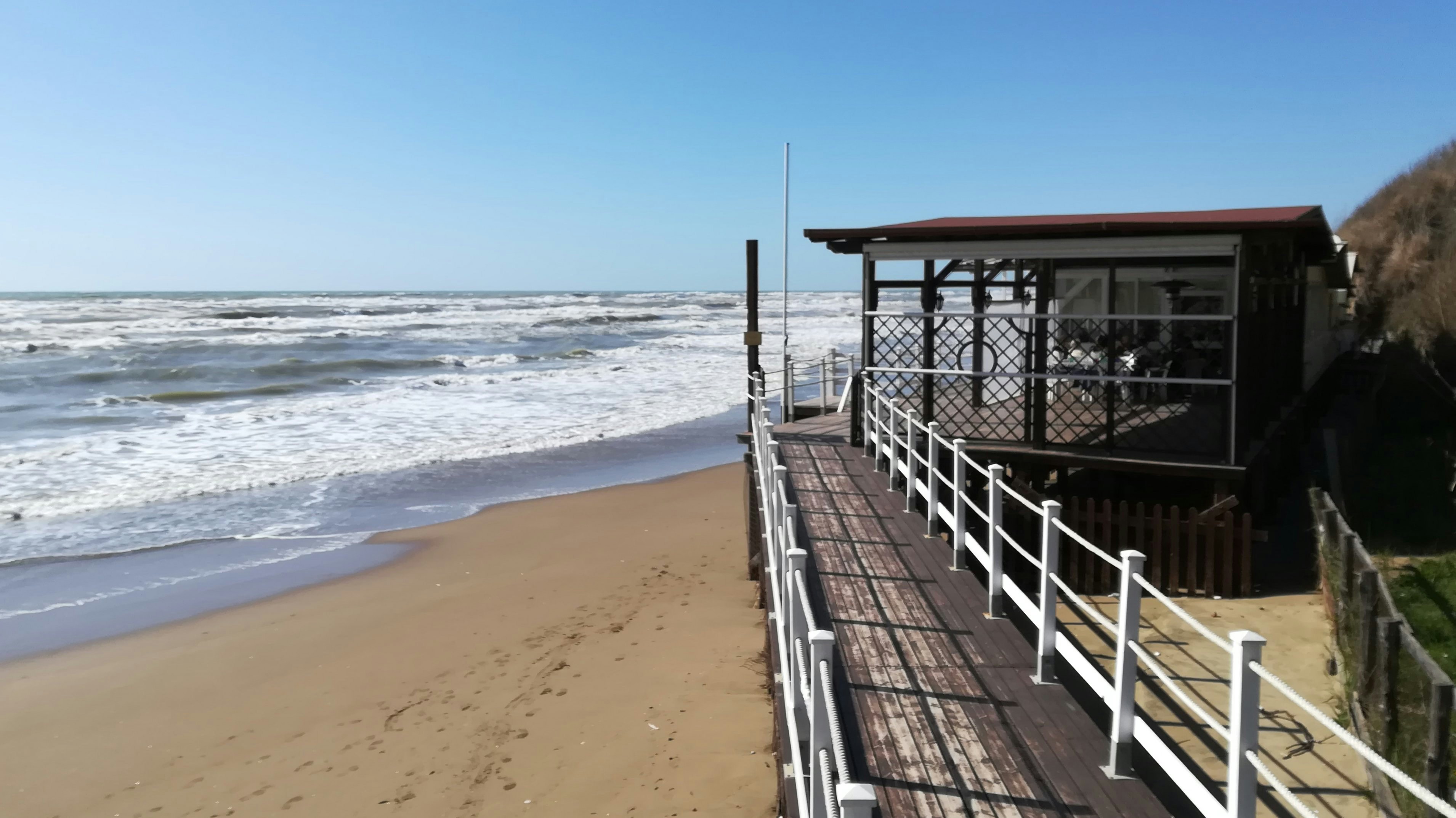 Wooden boardwalk leading to a seaside pavilion with waves crashing onto a sandy shore under a clear blue sky.