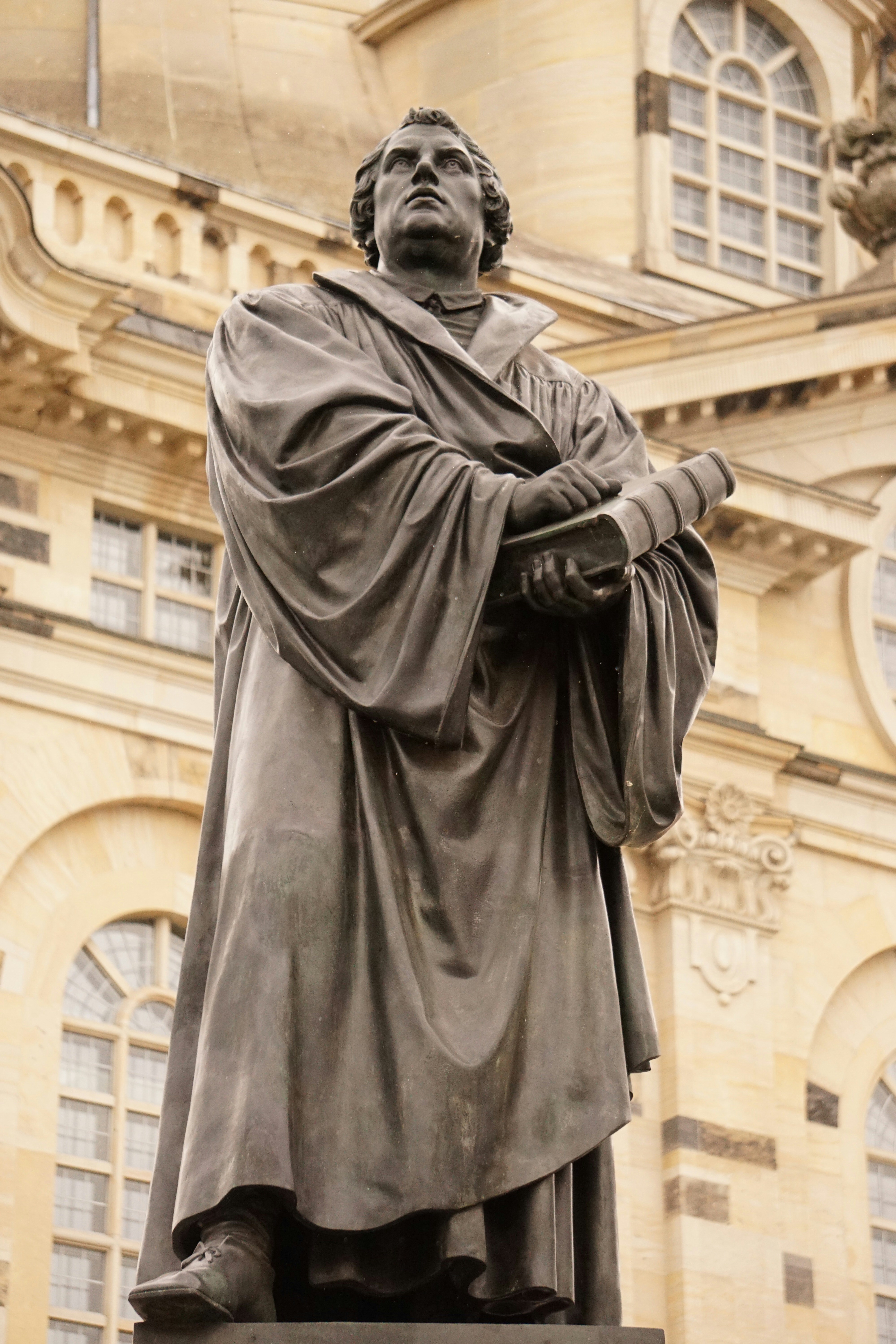 Bronze statue of a historical figure, draped in robes, gazing upwards with a scroll in hand, set against a grand architectural backdrop.