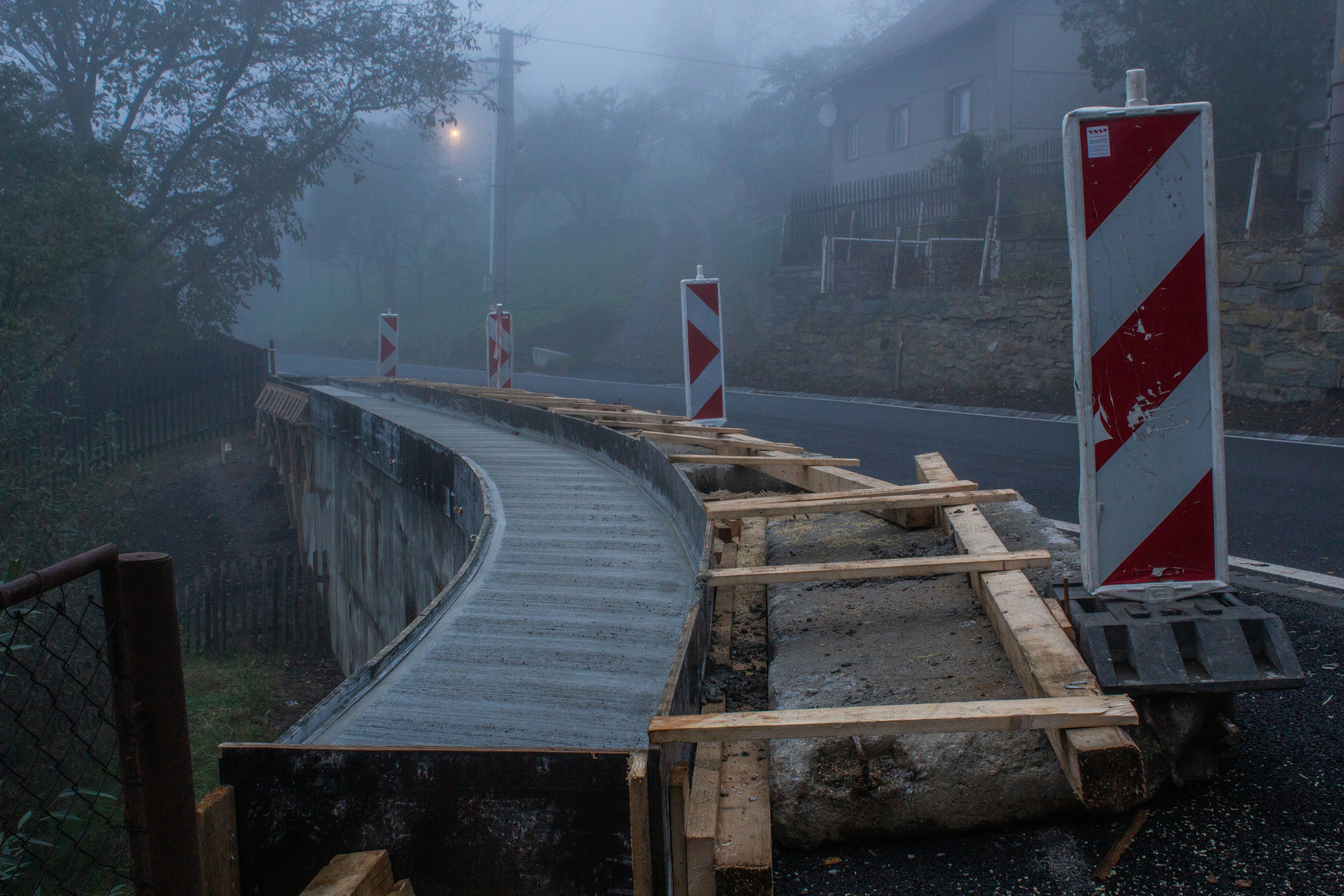 Curved road with construction barriers and fog enveloping nearby trees and structures.