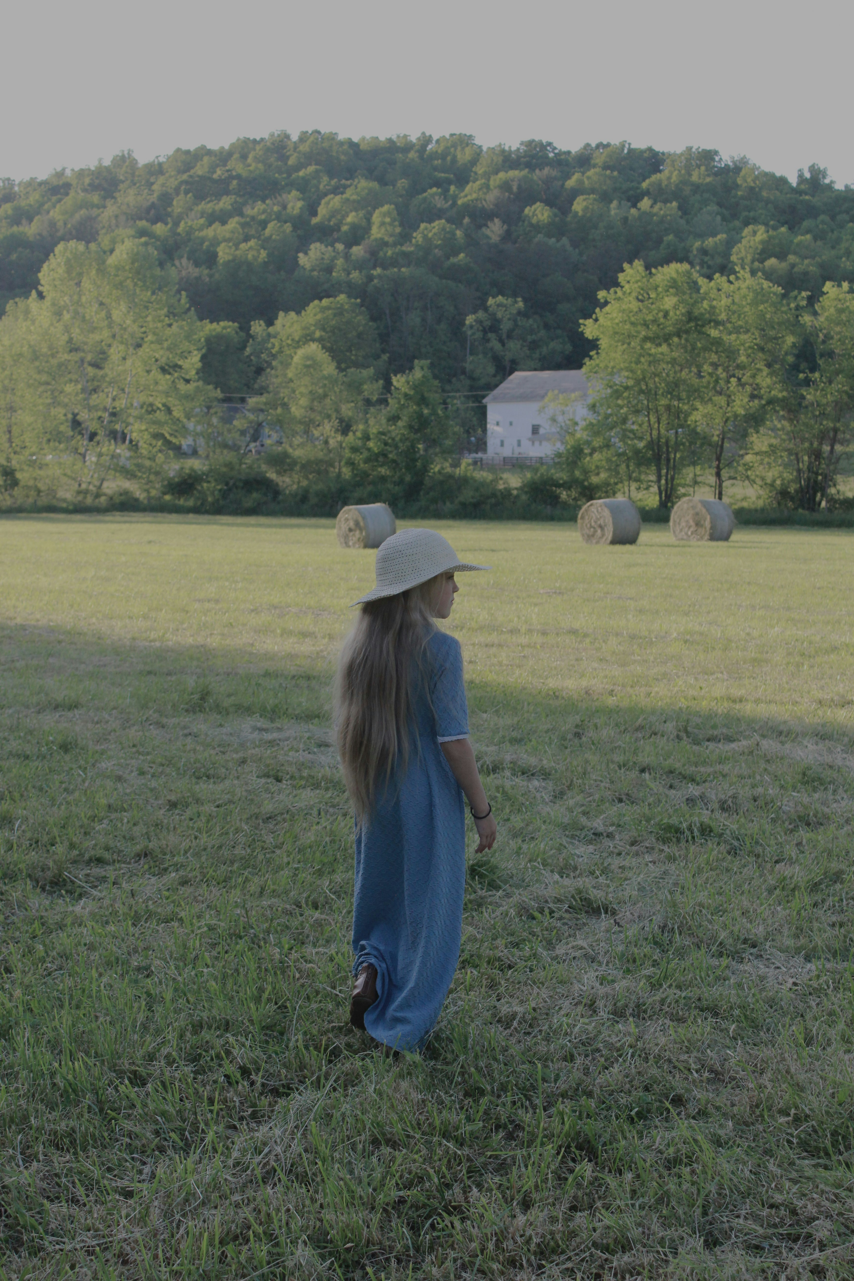 A young girl in a blue dress strolls through a sunlit field, her long hair cascading down her back, with hay bales dotting the landscape in the background.