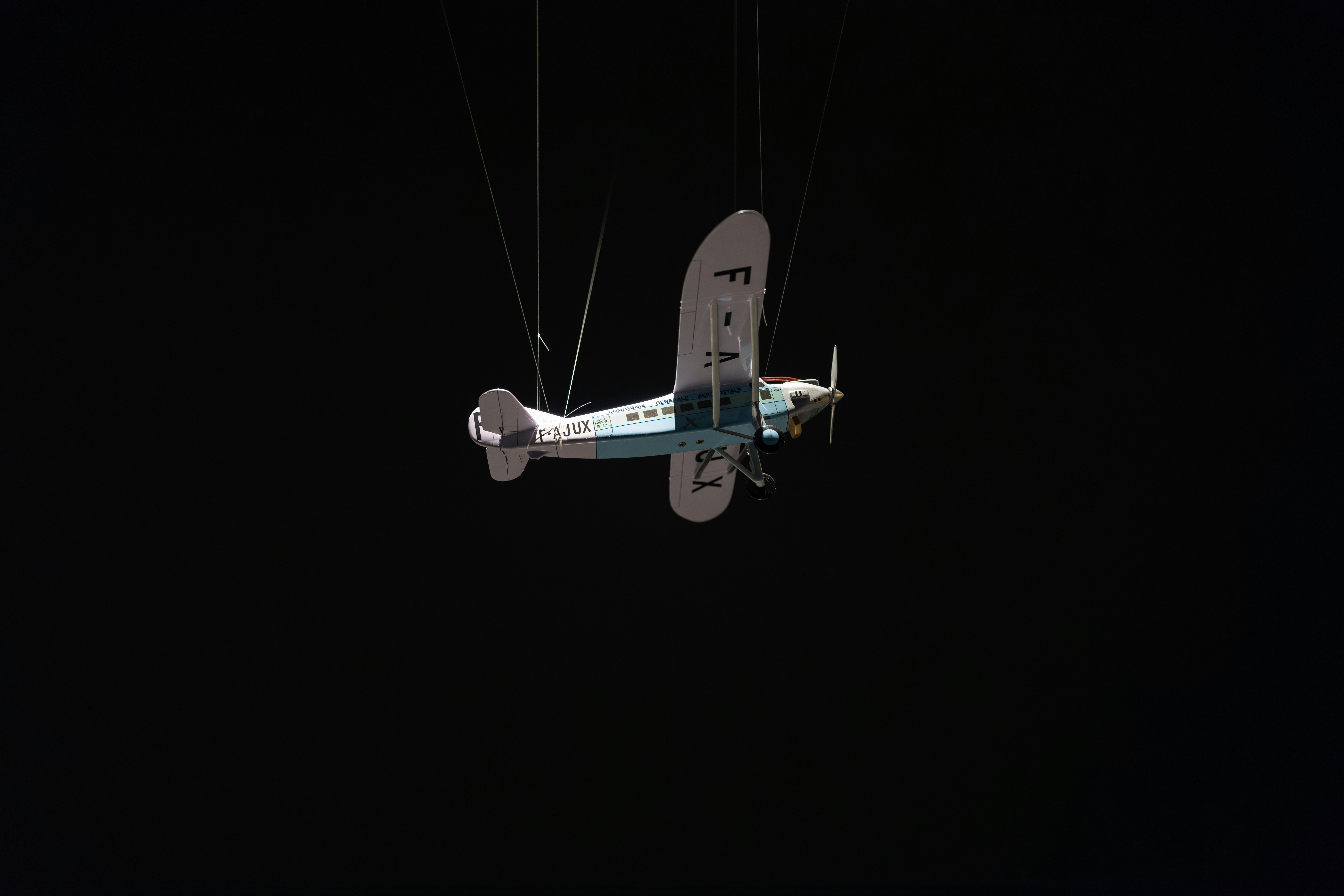 A small blue-and-white aircraft suspended by thin rigging lines against a black night, illuminated to reveal its details.