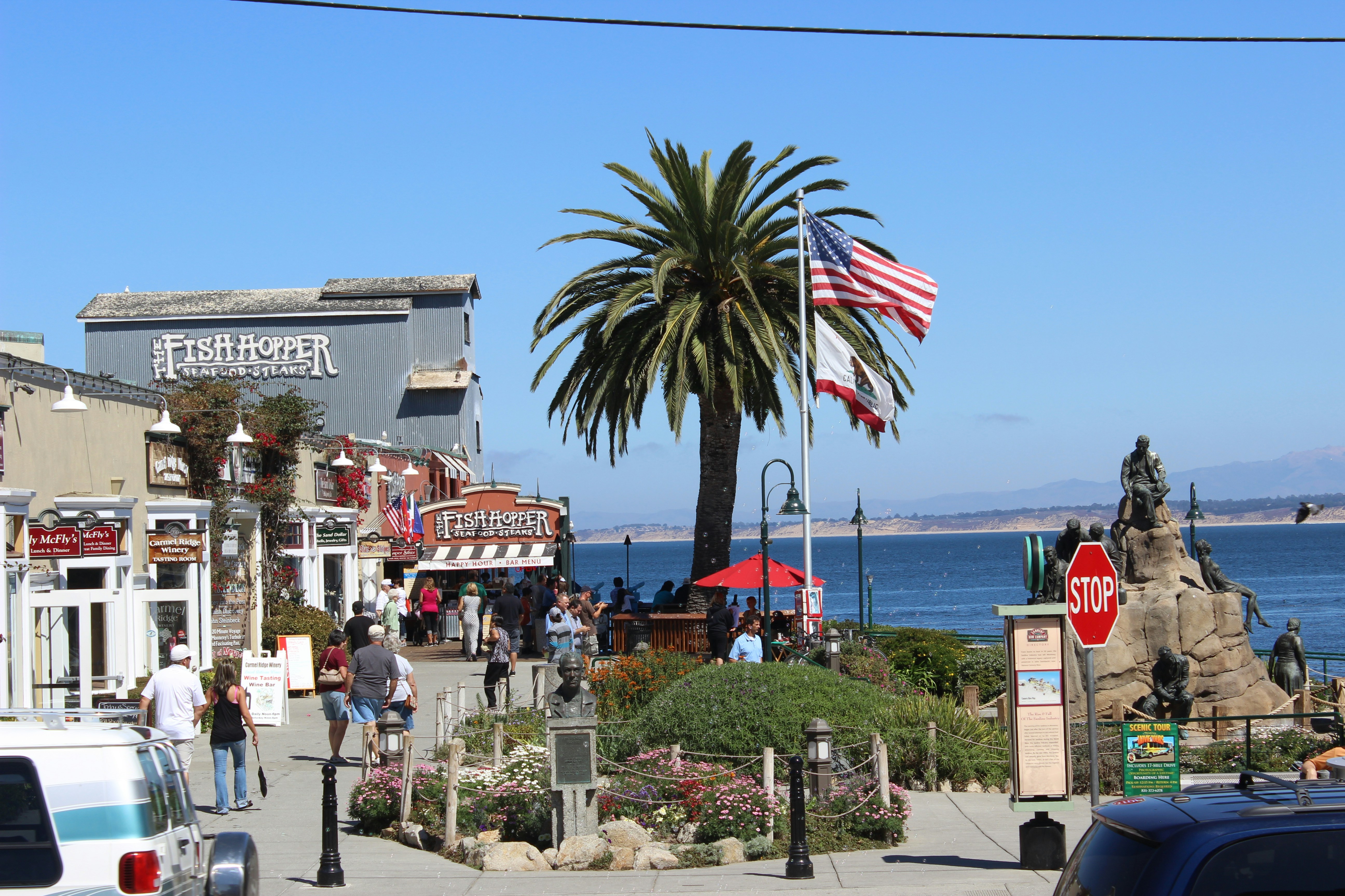 A palm tree and American flags line a bustling seaside promenade under a clear blue sky.