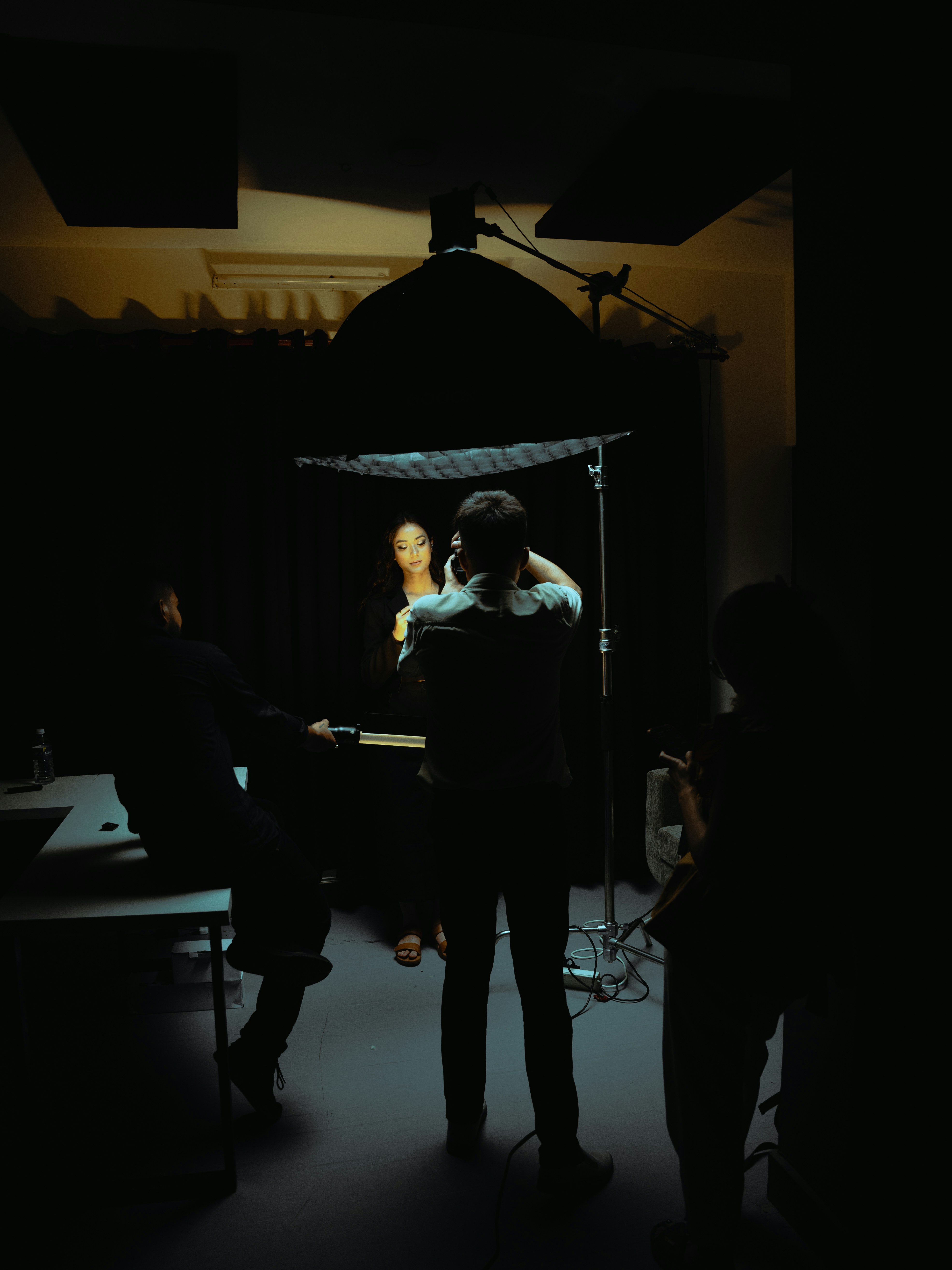 A focused photography session capturing a model under soft lighting, surrounded by a team adjusting equipment. The contrast of light and shadow enhances the atmosphere.
