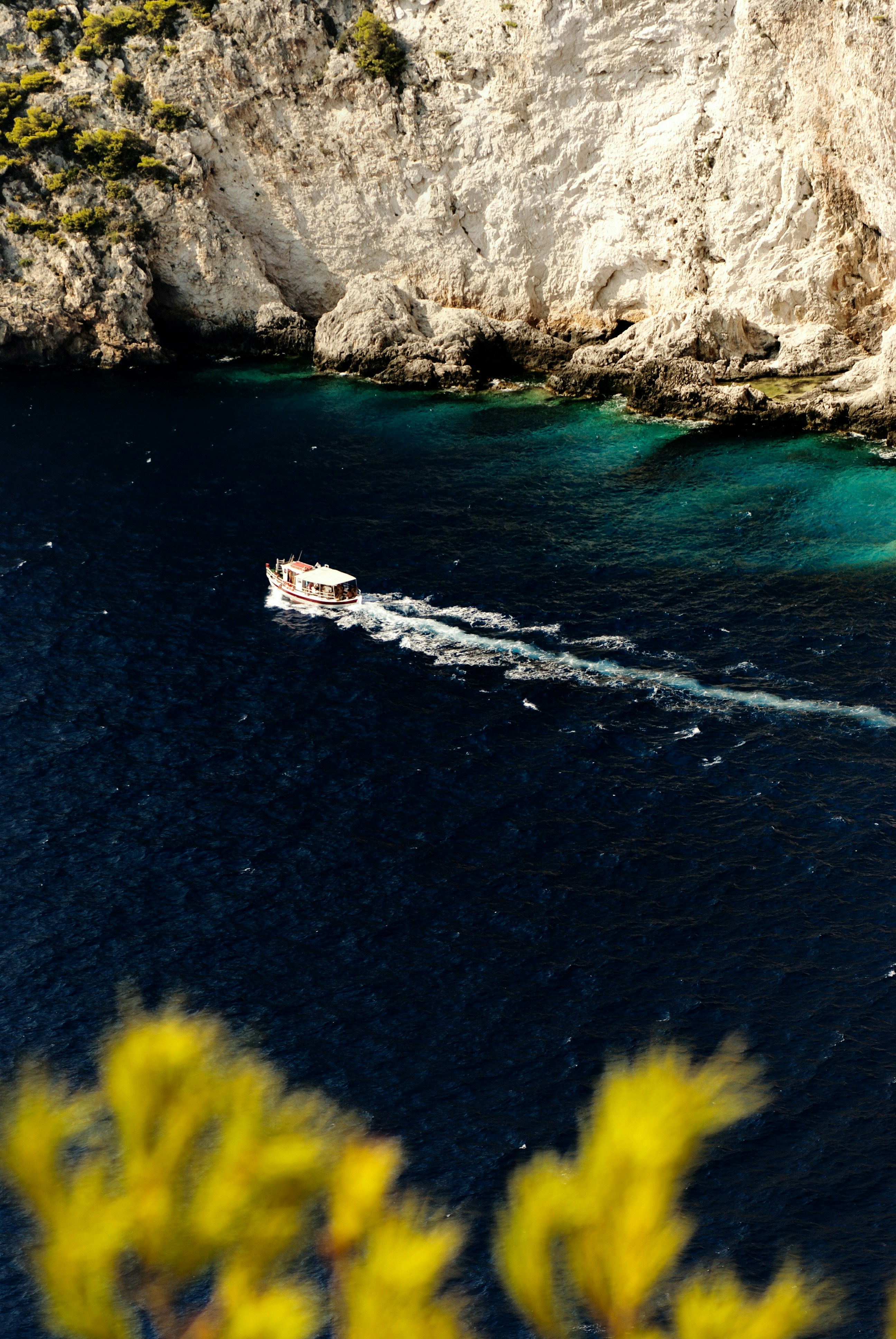 Small motorboat glides across deep blue water beside rugged white cliffs, where turquoise shallows meet the rocky shore.